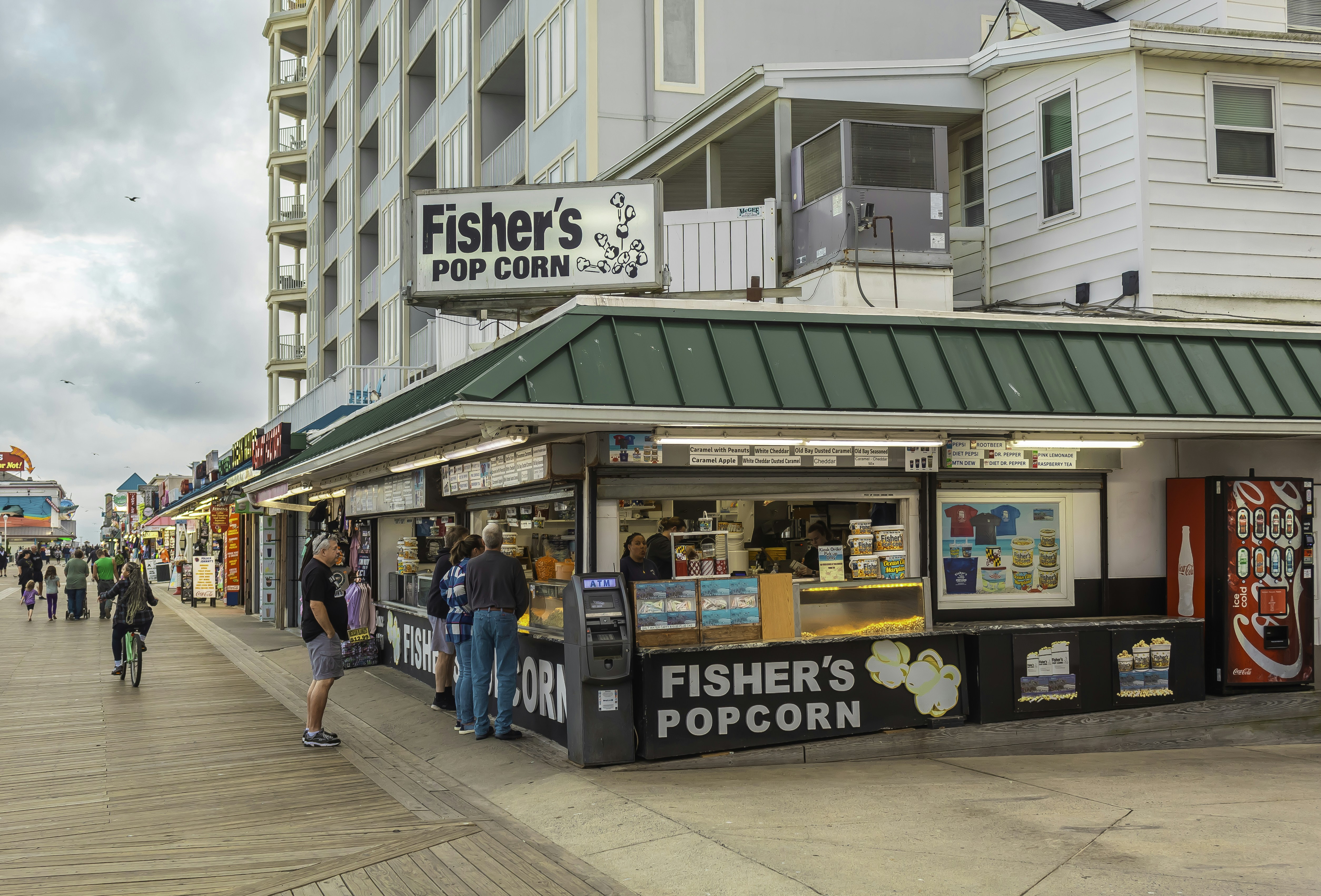 Popcorn stand on a bustling boardwalk with people queuing, set against pastel buildings under an overcast sky.