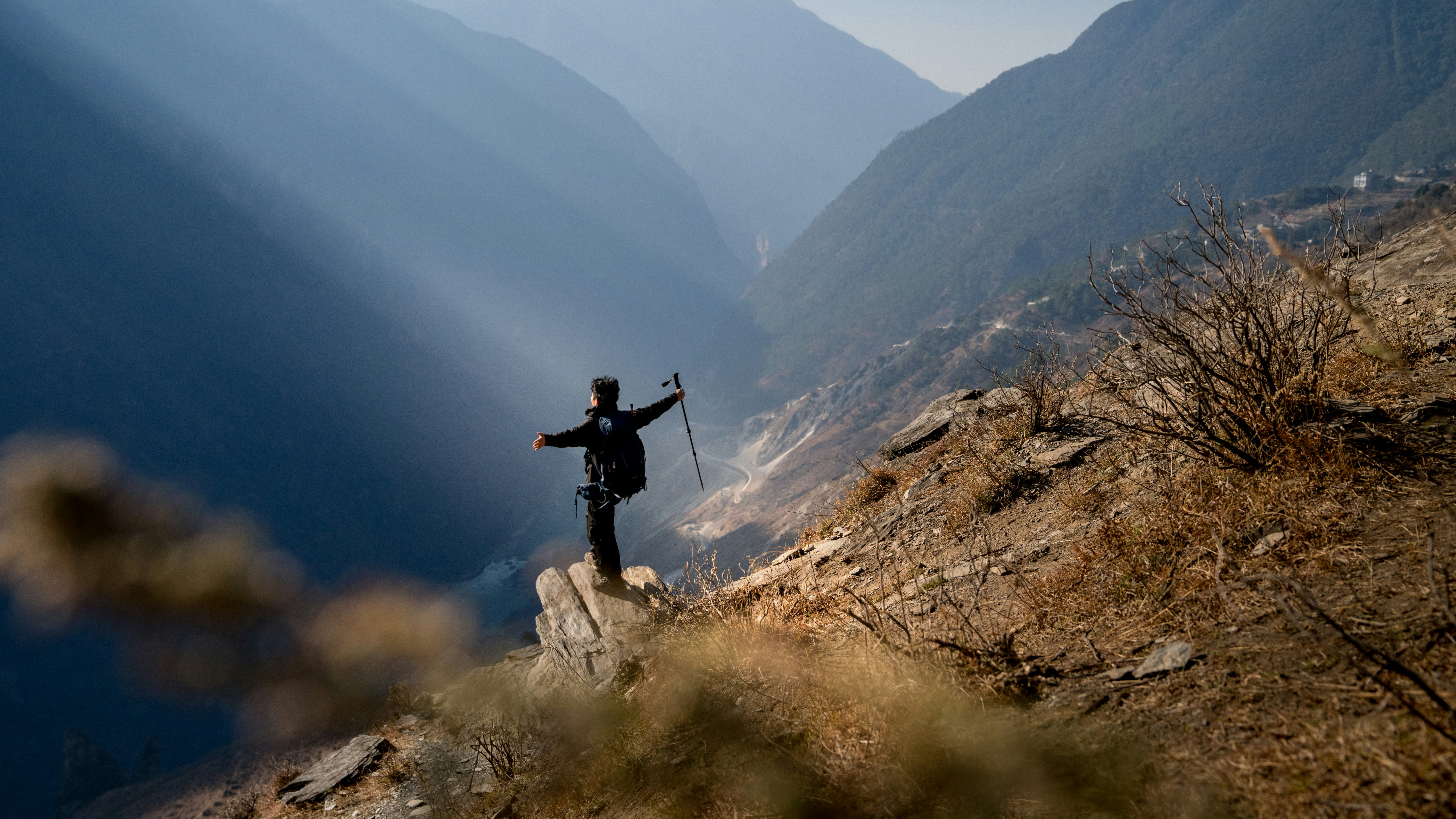 Hiker standing on a rocky outcrop with arms outstretched, bathed in sunlight streaming through a mountainous valley.