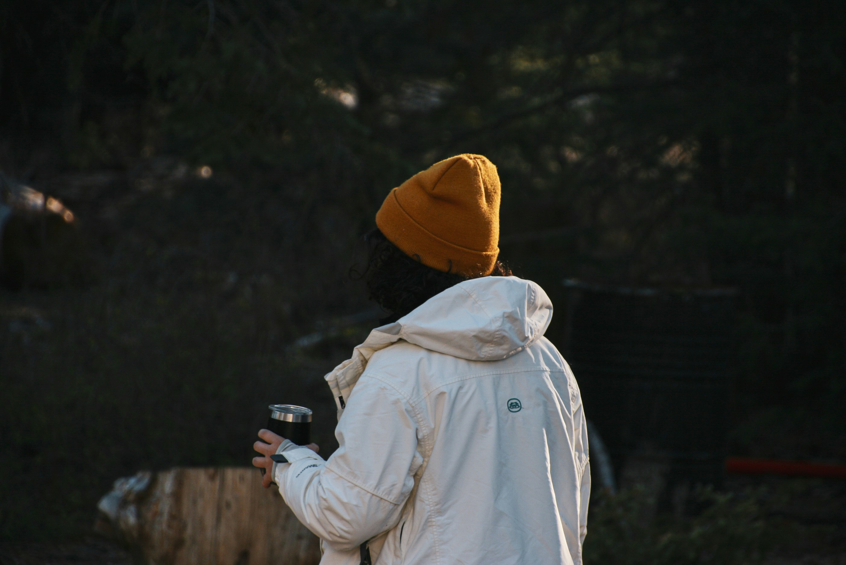 Person in a white jacket and orange beanie holding a mug, standing in a forest clearing.