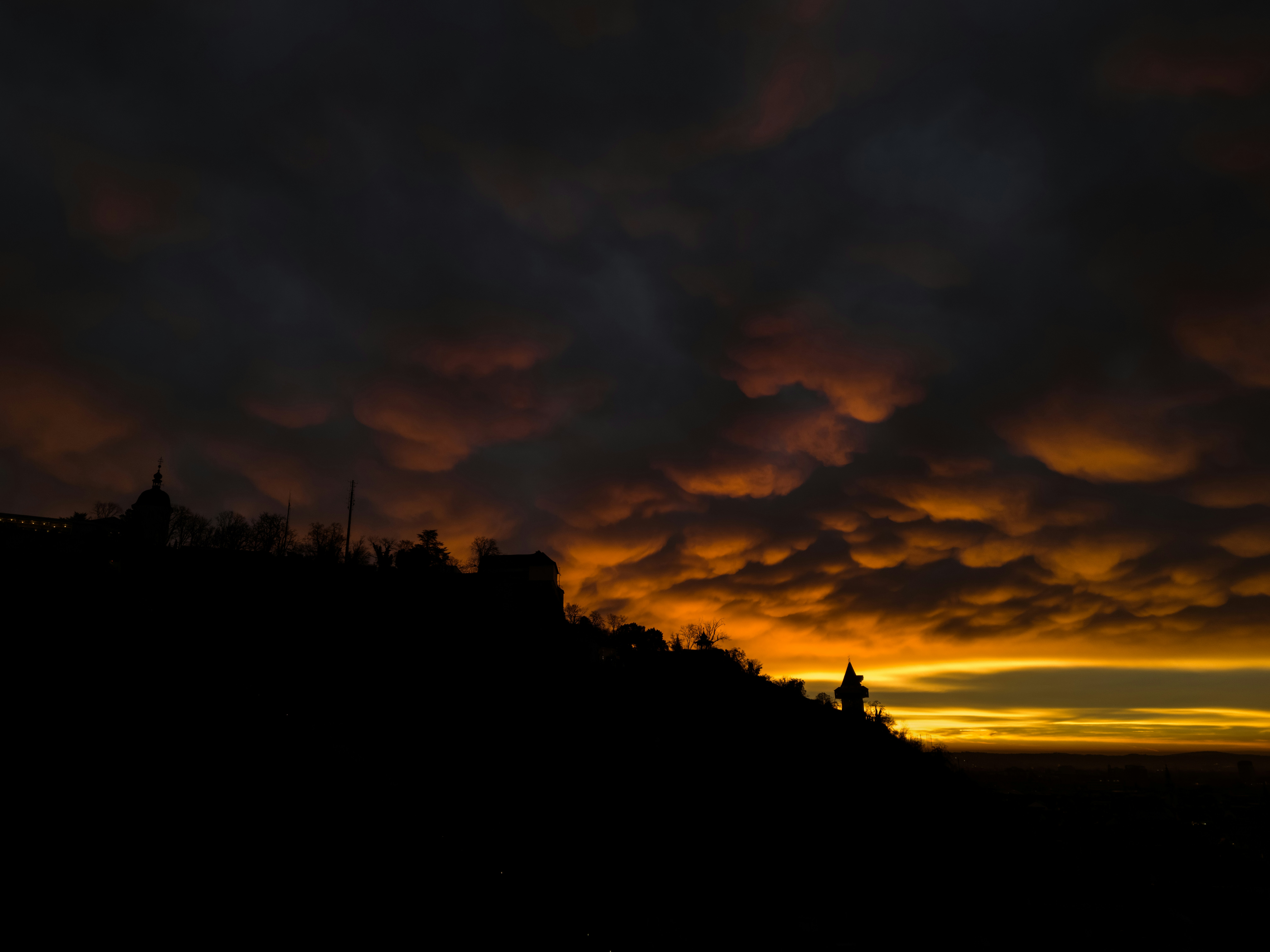 Dark clouds illuminated by vibrant oranges and yellows of a sunset over silhouetted hills.