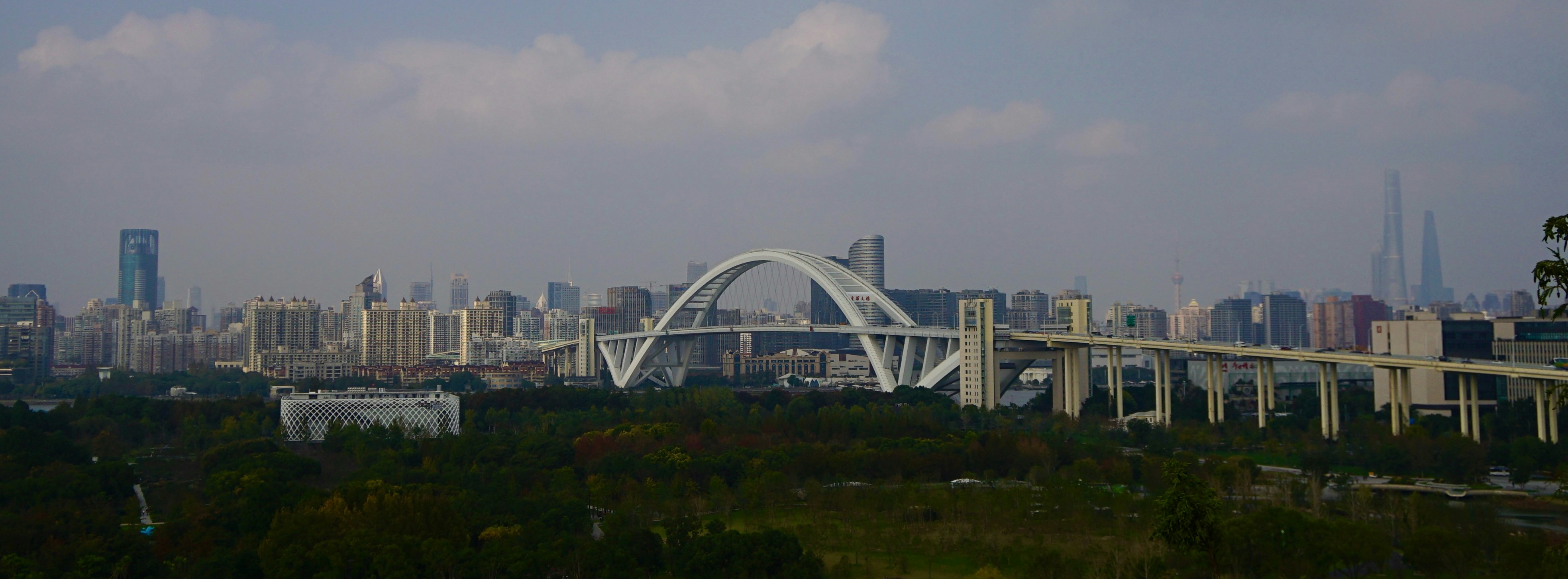White arched bridge spans a vibrant city skyline under a cloudy sky.