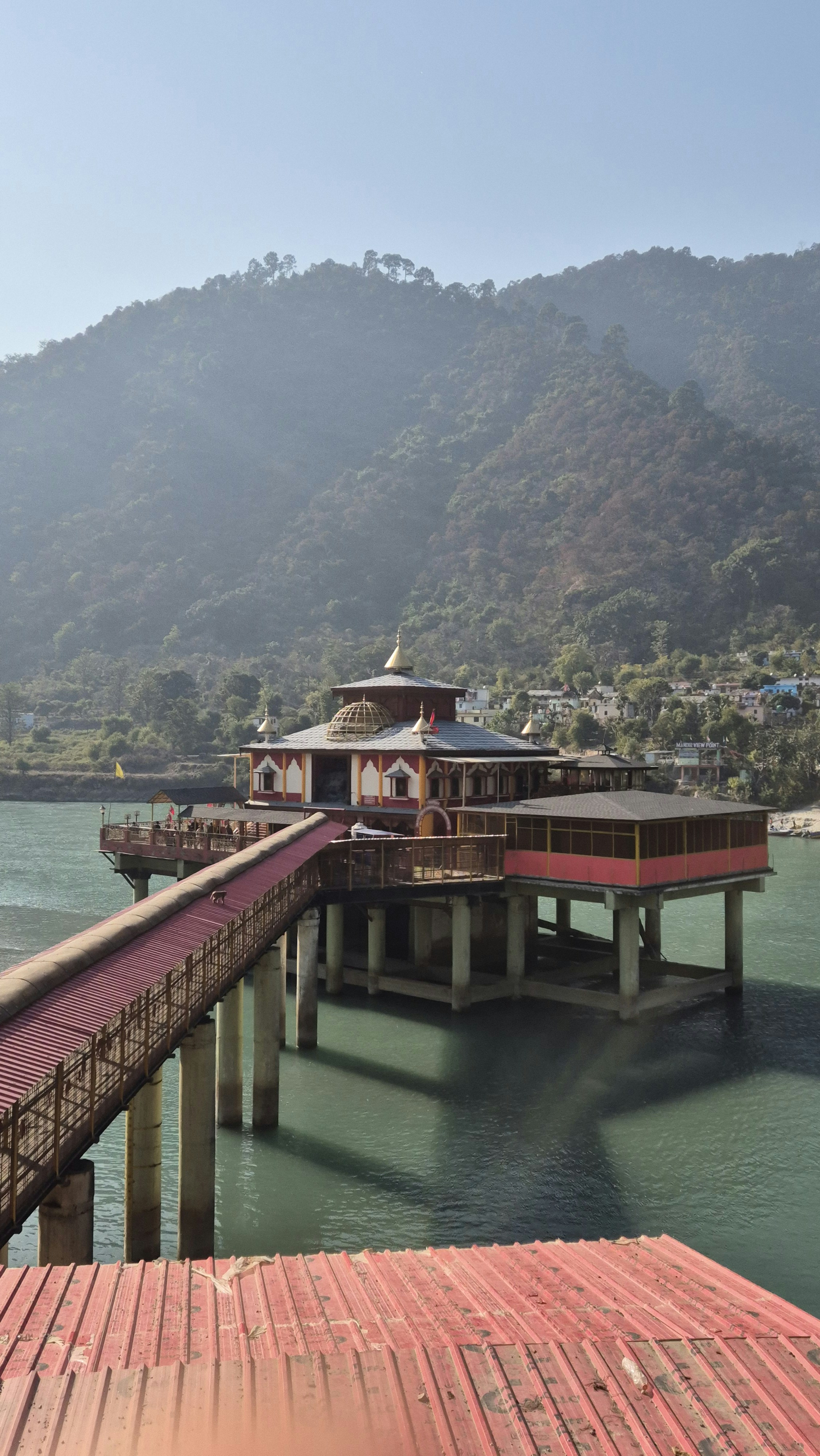 Temple-style building on stilts rises over a calm lake, reached by a long wooden walkway. Forested hills rise in the background under a clear sky.