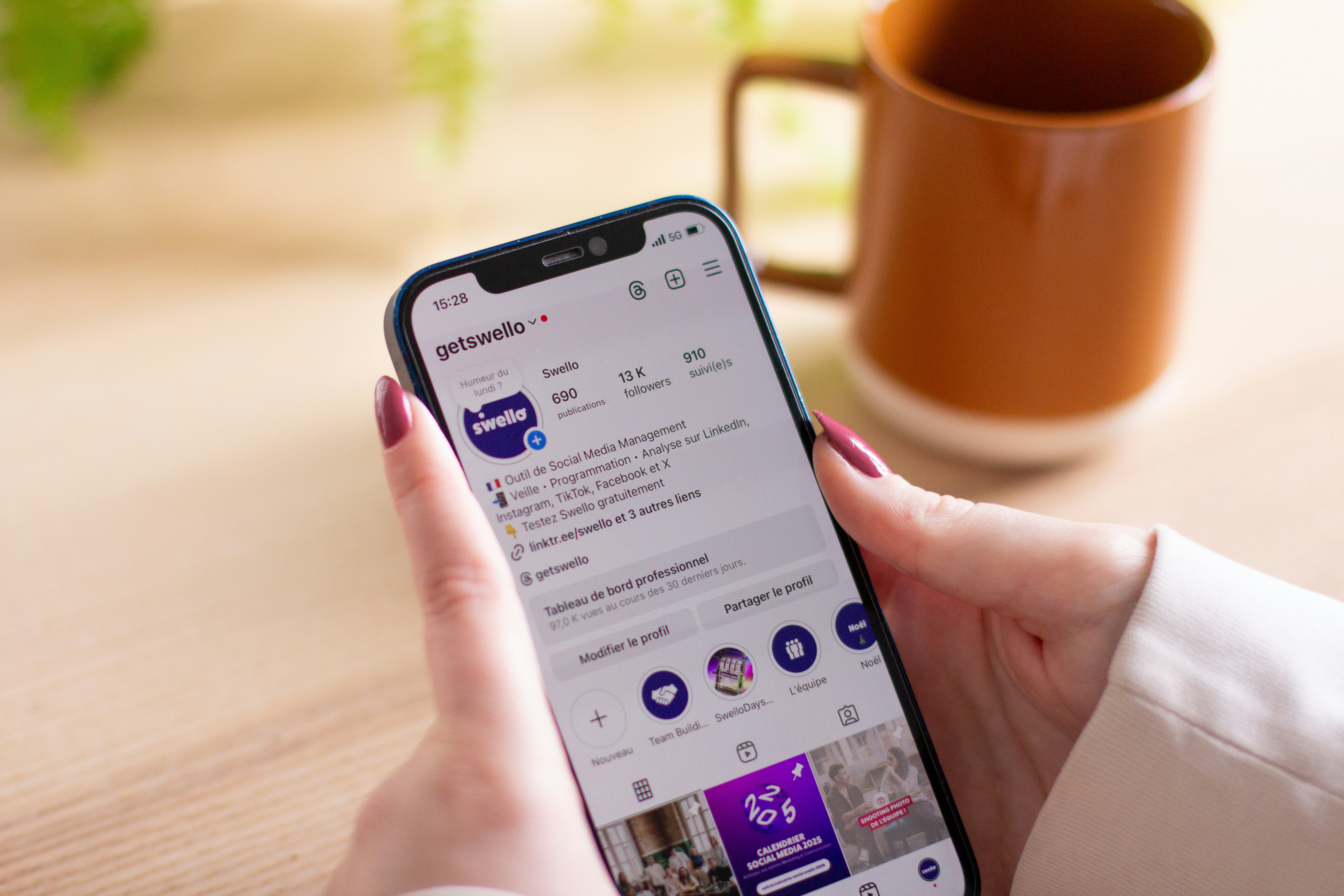 Hands holding a smartphone displaying a social media profile, with a blurred coffee mug in the background.