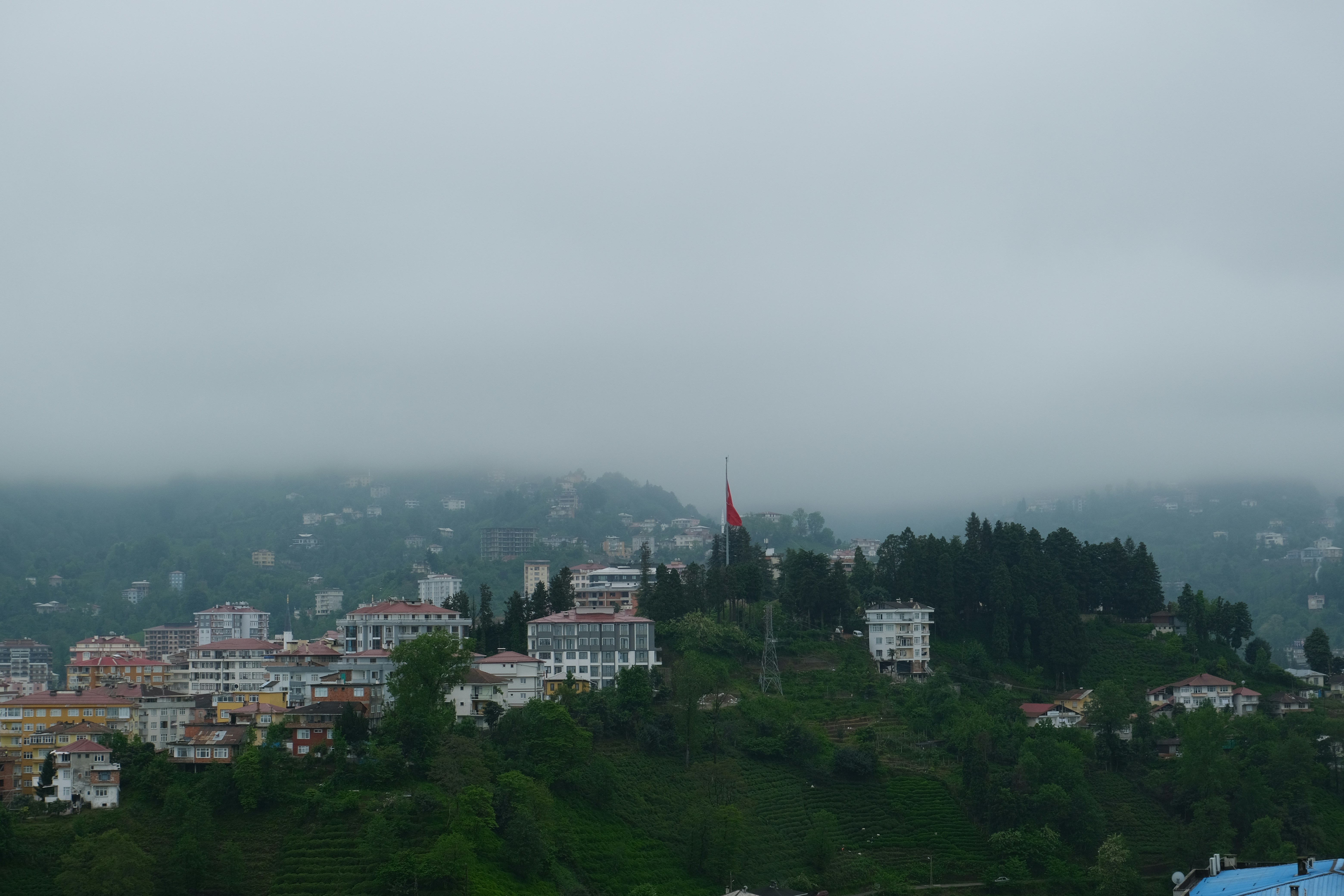 Clouds blanket a lush hillside dotted with buildings and a prominent flag under a gray sky.