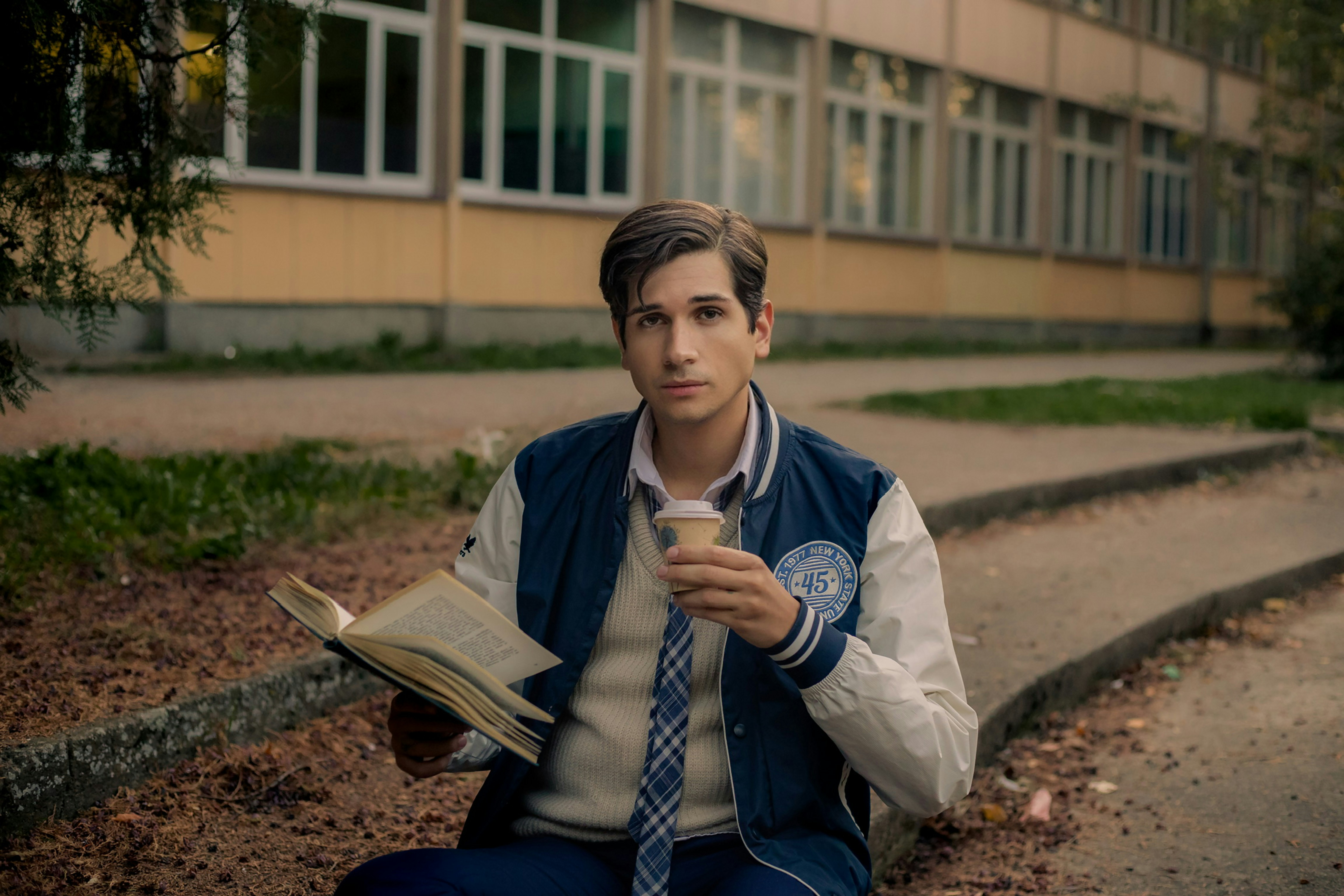 Person in a varsity jacket holding a book and drinking tea while seated near a school building.