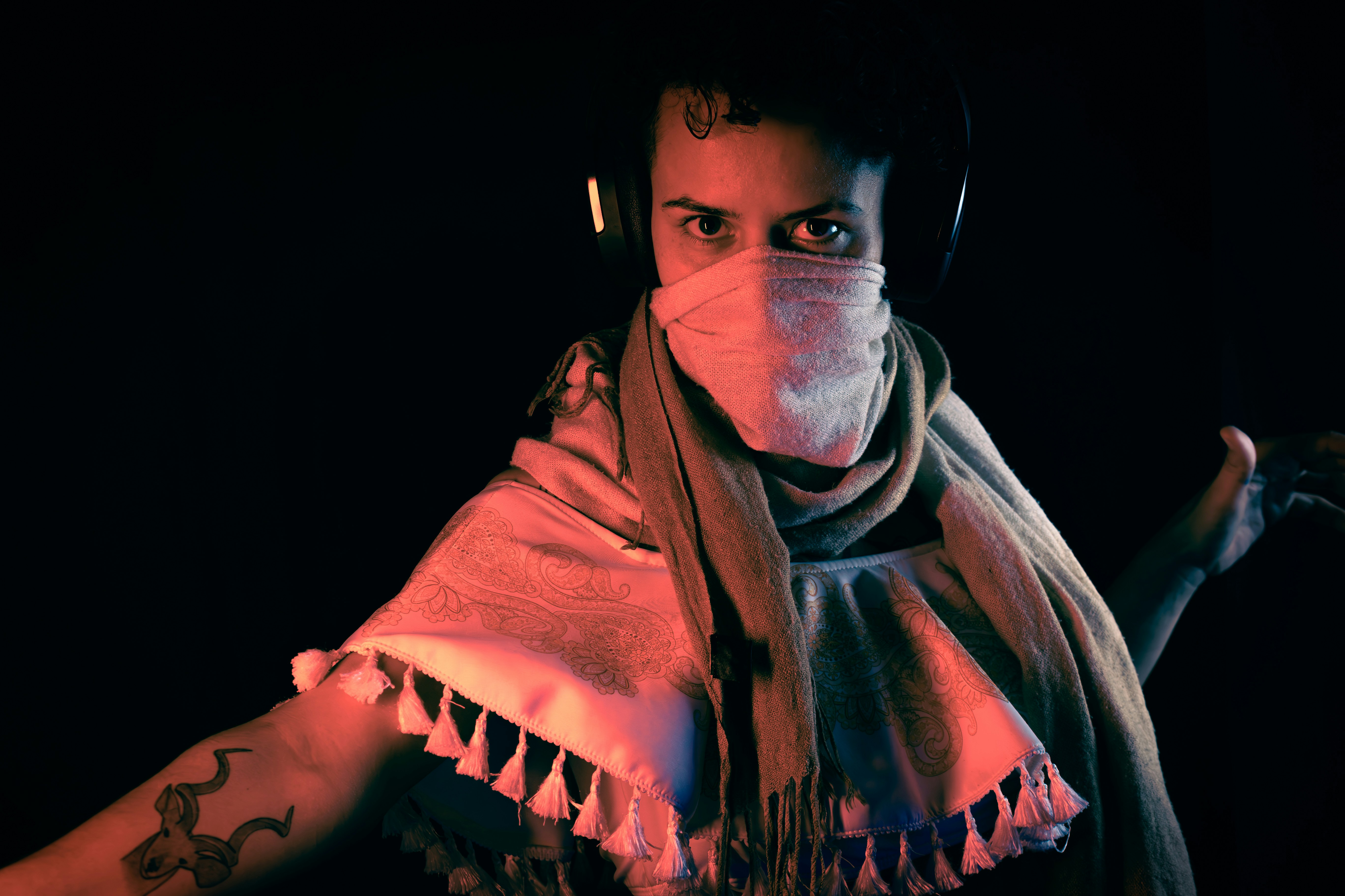 Person in a scarf and headphones, face partially covered, stands with arms slightly raised against a dark background.