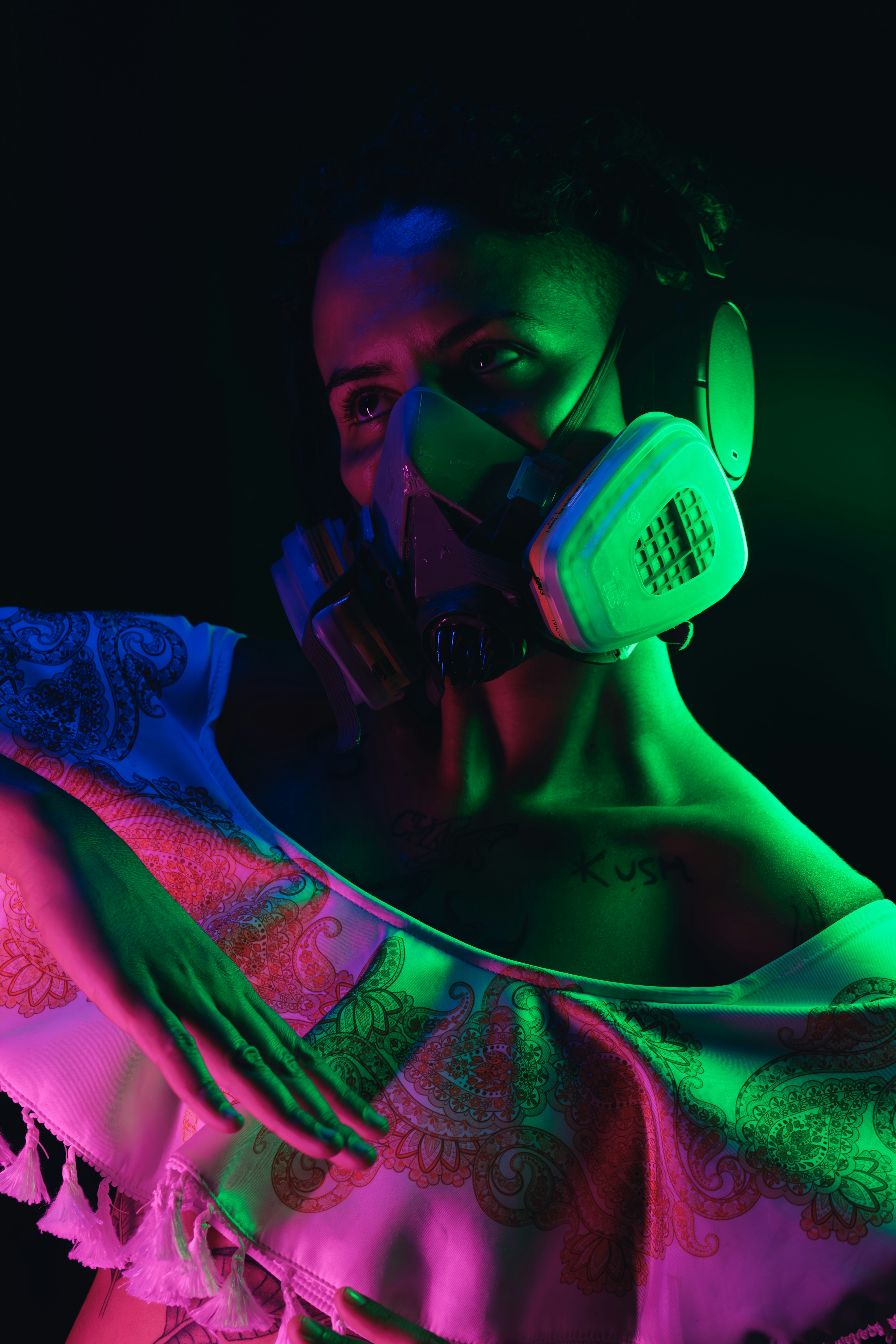 A figure in a respirator showcases intricate fabric draping, illuminated by vibrant green and purple lighting.