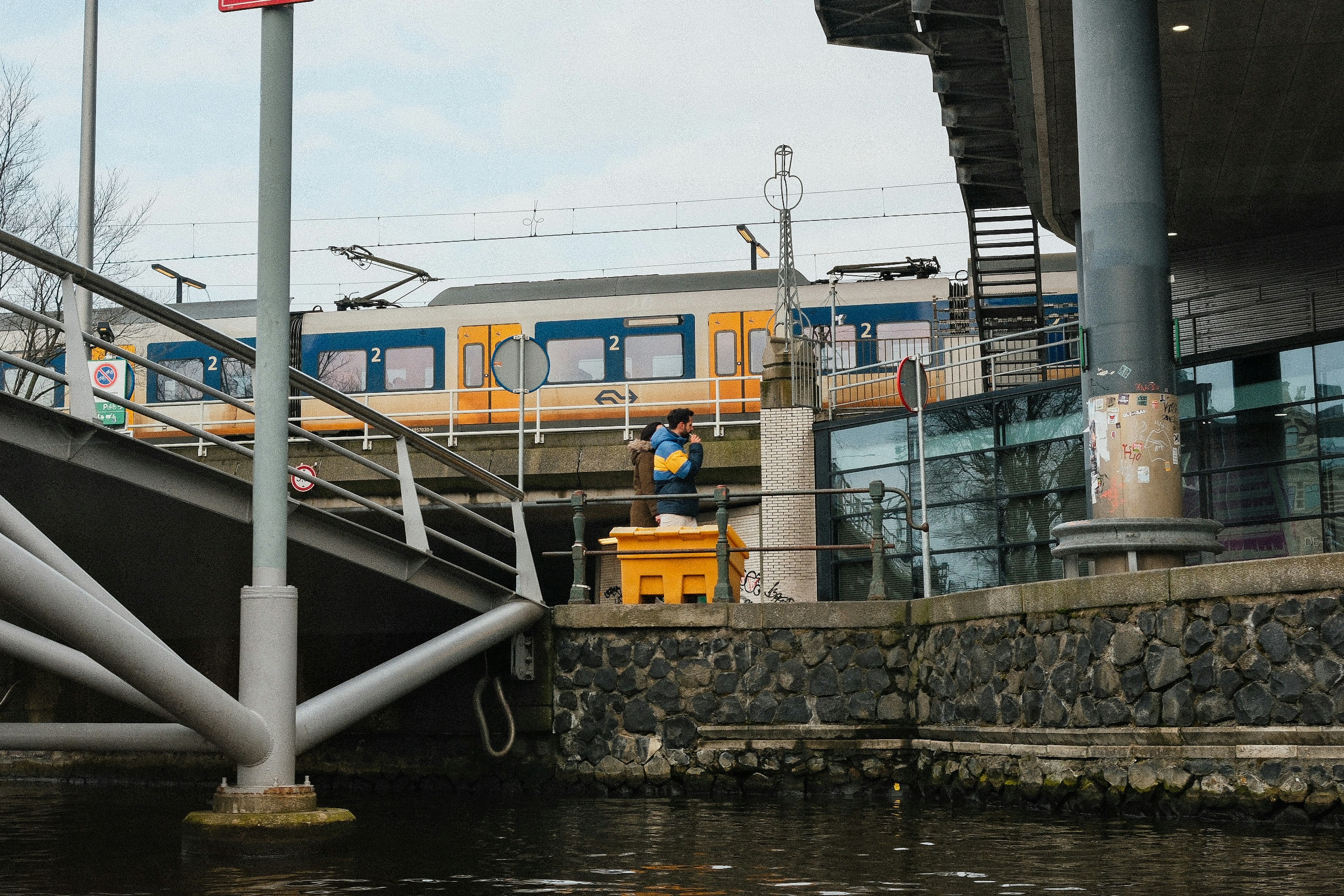 Man standing near a canal under a railway bridge with a yellow train passing above.