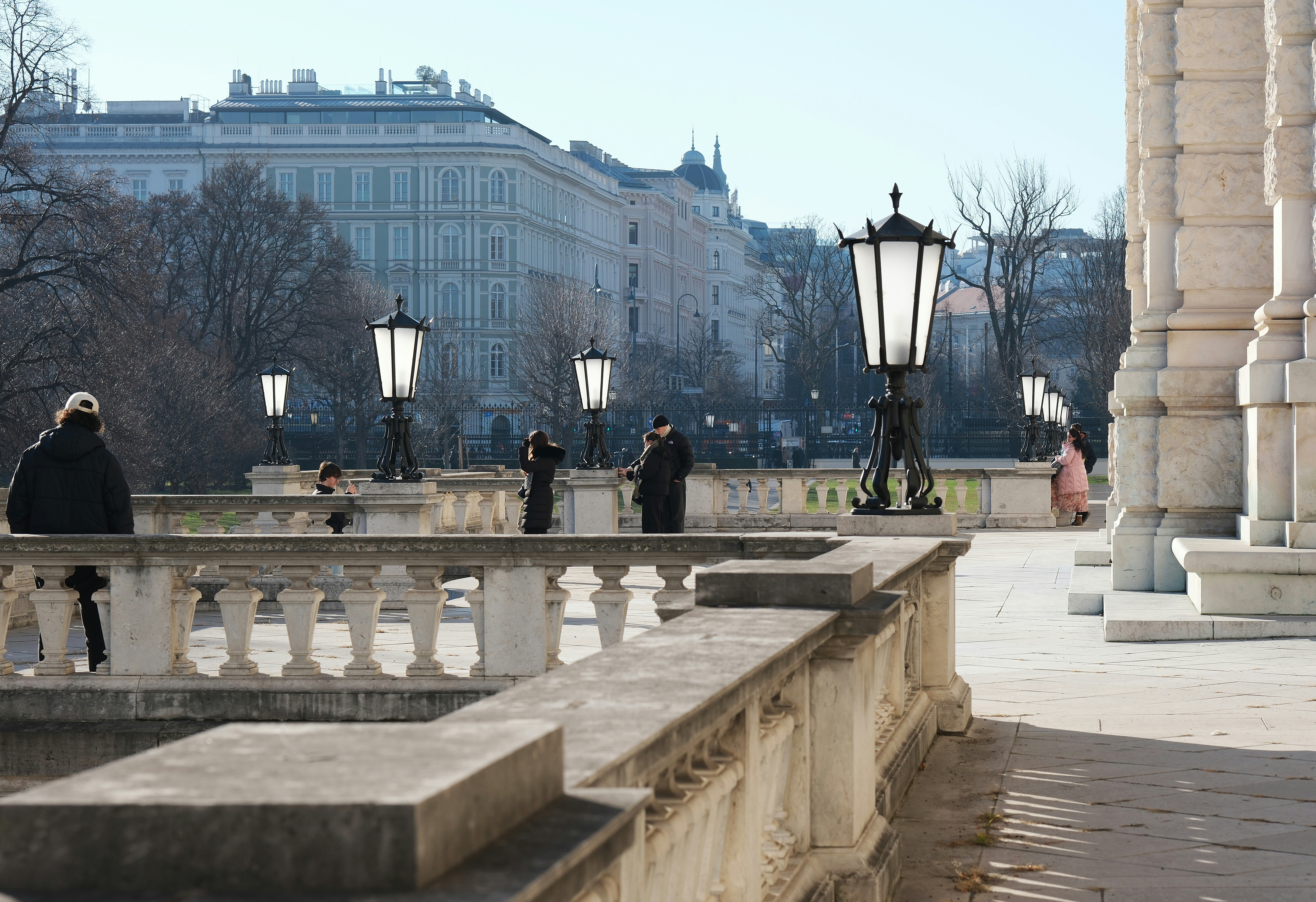 People walking along a stone balustrade with ornate lanterns under a clear blue sky.