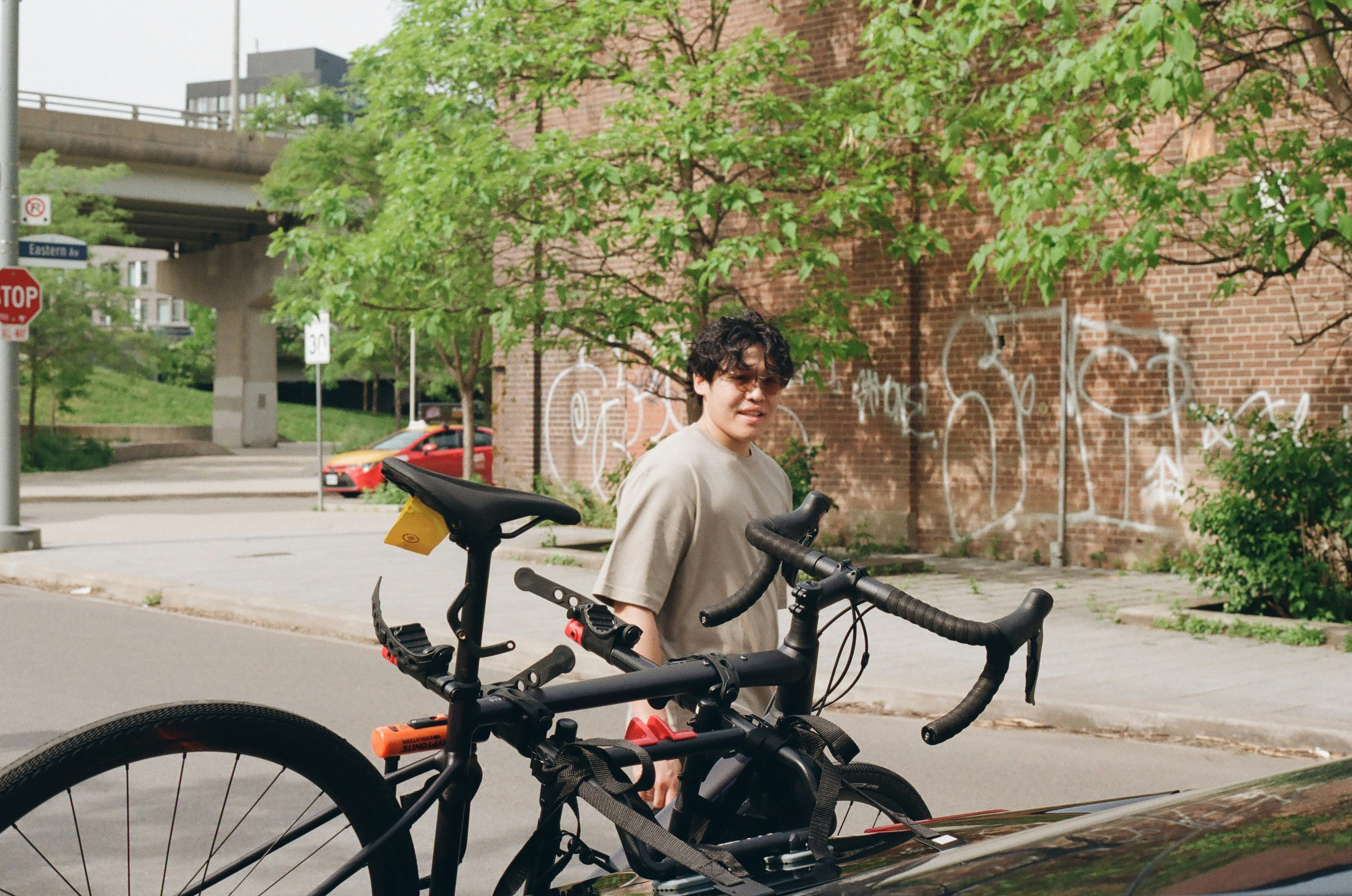 A candid photograph capturing a young man with a road bike paused beside a graffiti-covered brick wall along a quiet urban street. The scene emphasizes urban texture and the momentary pause in a city rhythm.
