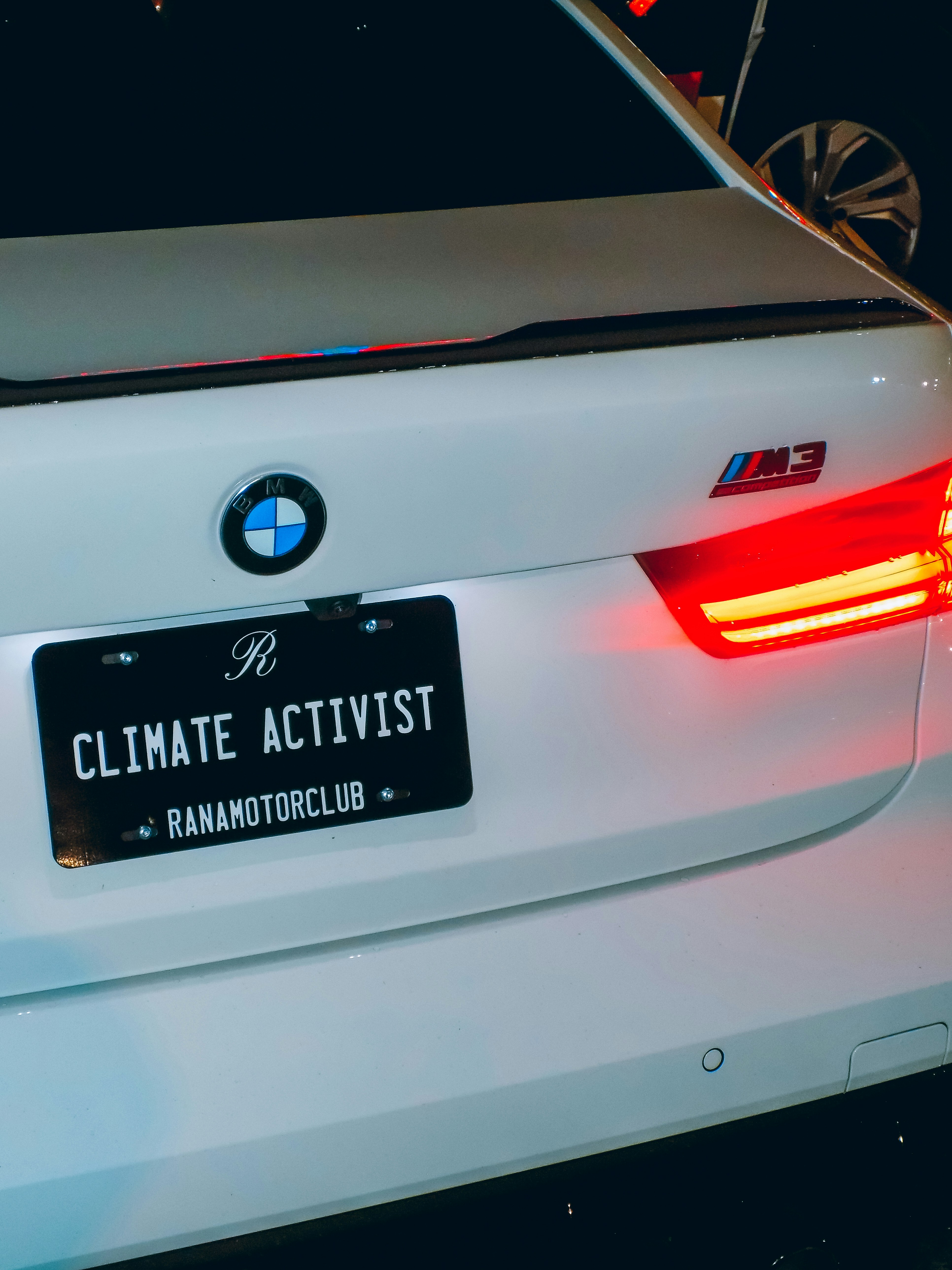 Rear view of a white BMW showing a license plate reading 'CLIMATE ACTIVIST' beside a bright red taillight. The shot emphasizes the plate's message against the car's sleek M3 badge.