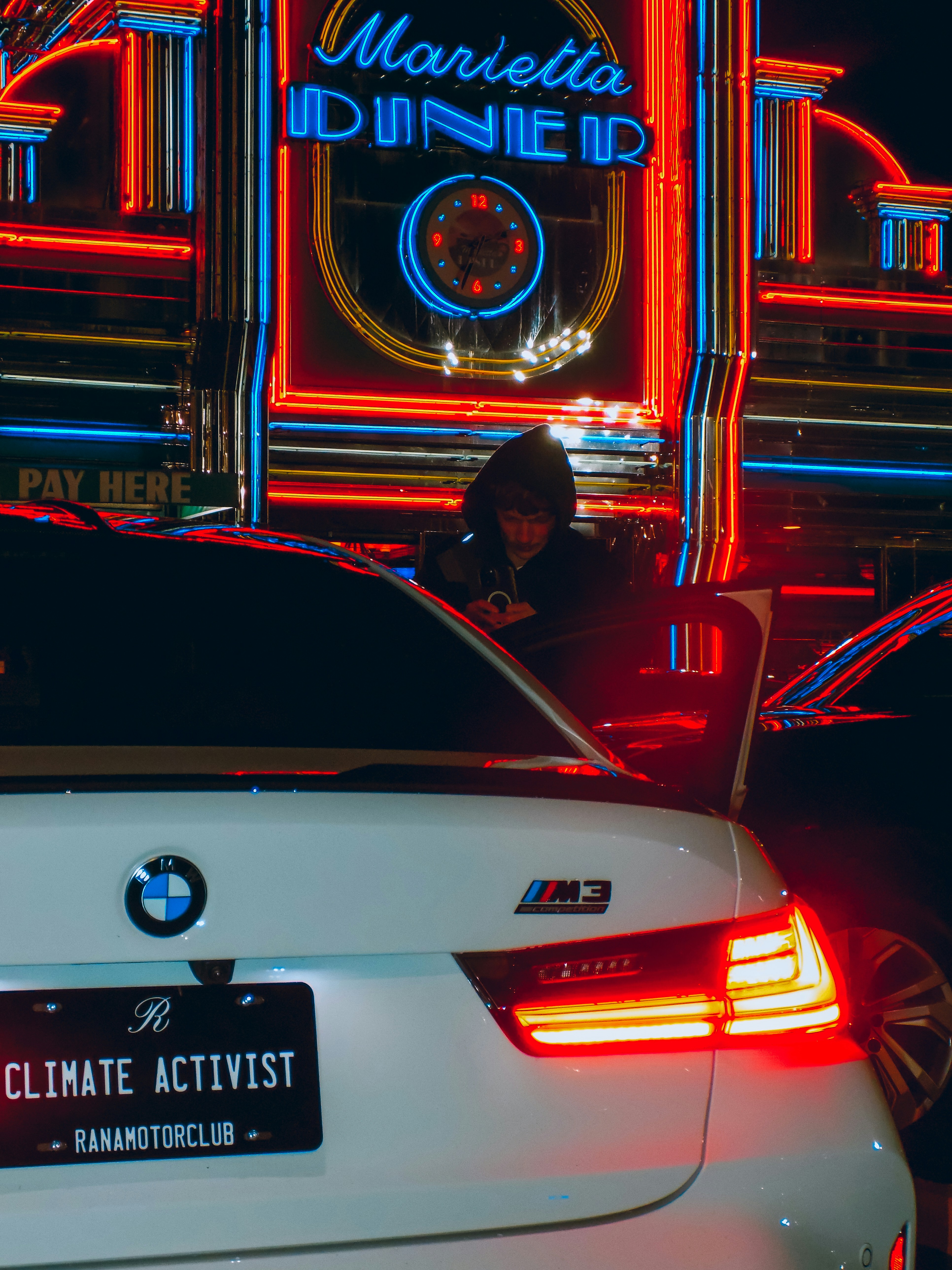 White BMW sedan with illuminated M3 badge sits under a neon-lit diner sign at night; a hooded figure stands beside the car, casting moody shadows as red and blue lights ripple across chrome and glass. This is a photograph with cinematic urban flair.