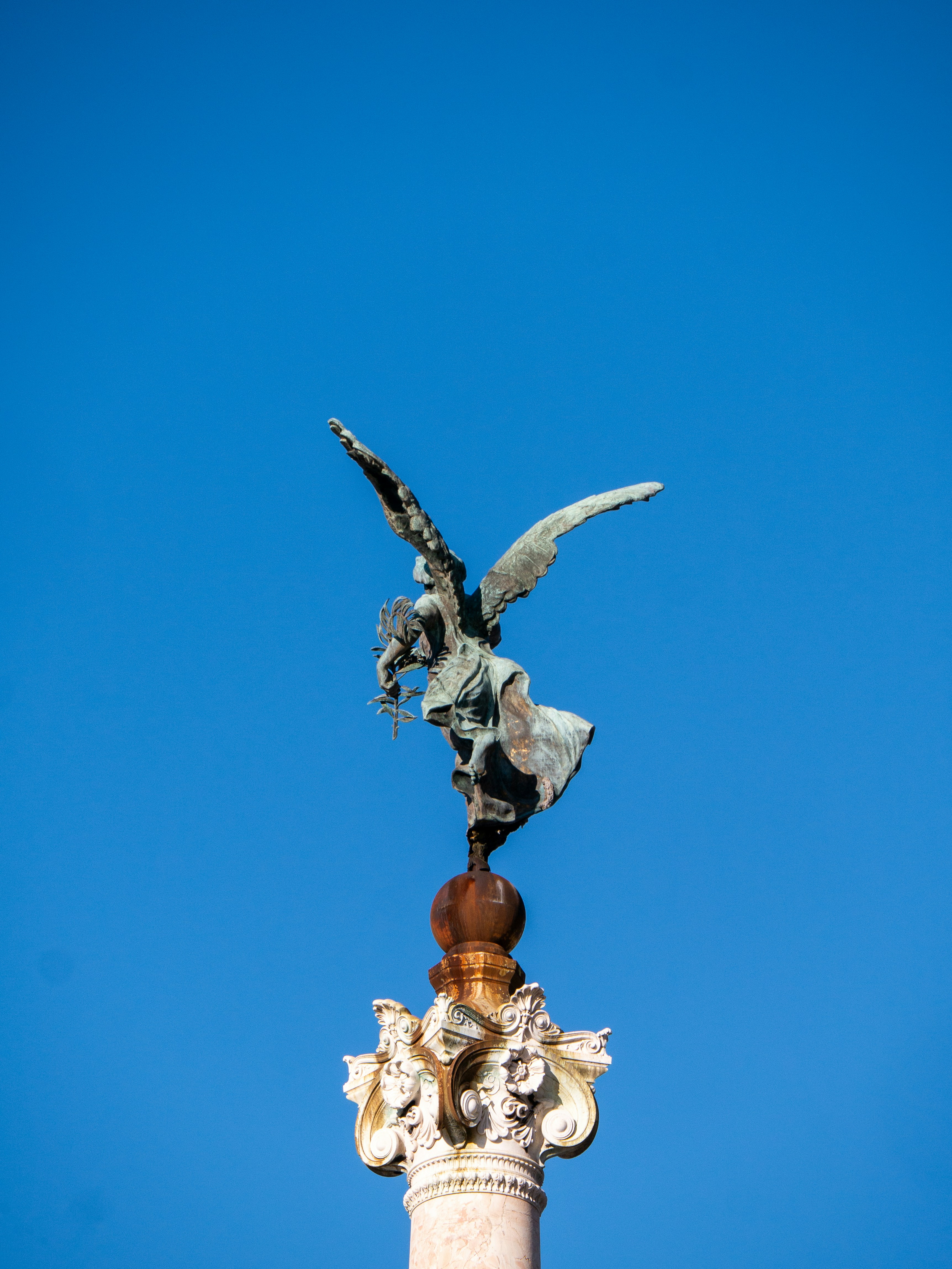Bronze angel sculpture perched atop a marble column against a clear blue sky.