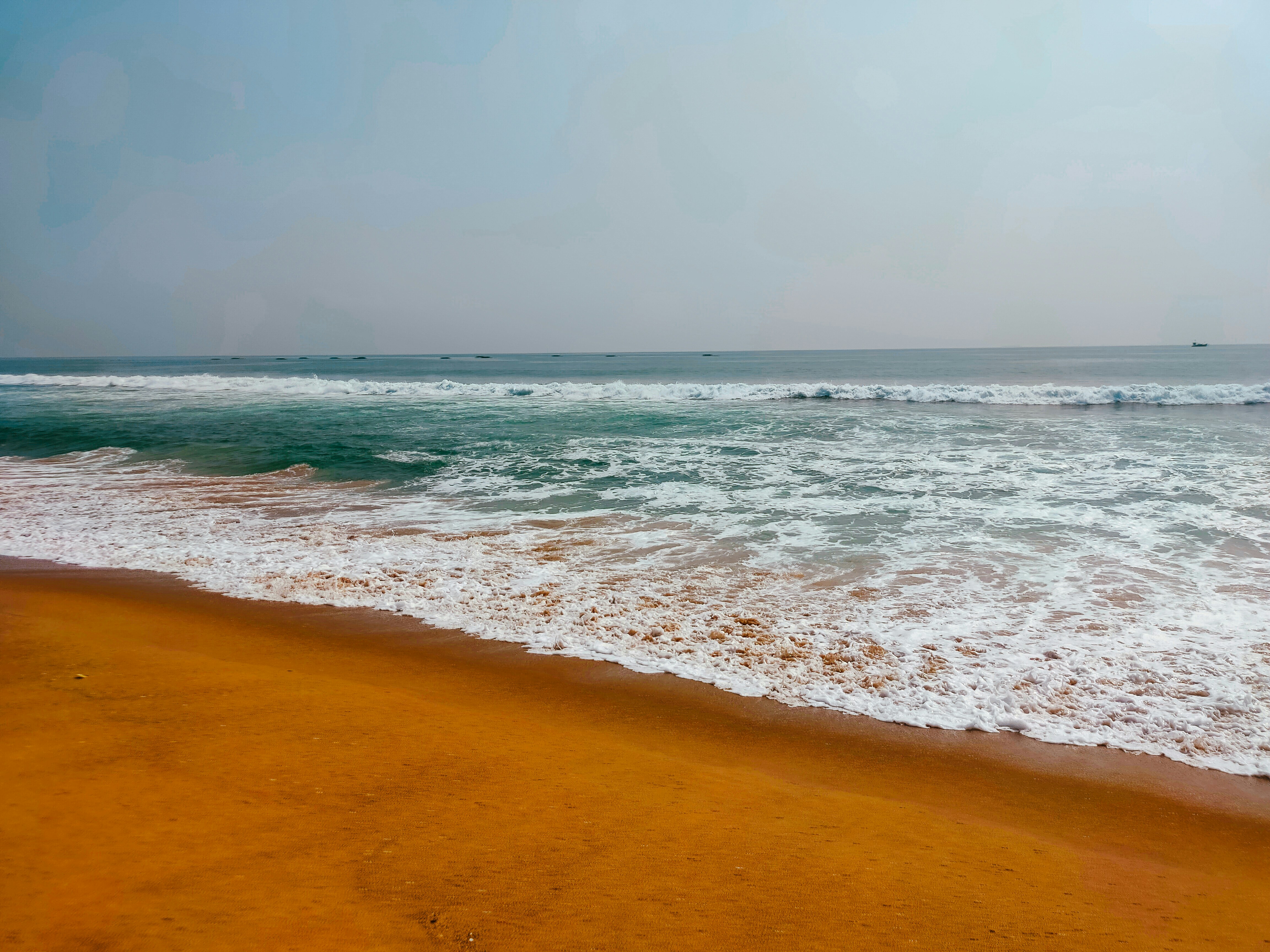 Sunlit beach with vivid orange sand meeting foamy turquoise waves beneath a pale blue sky. A wide coastal scene that emphasizes warm shoreline tones against the cooler sea.