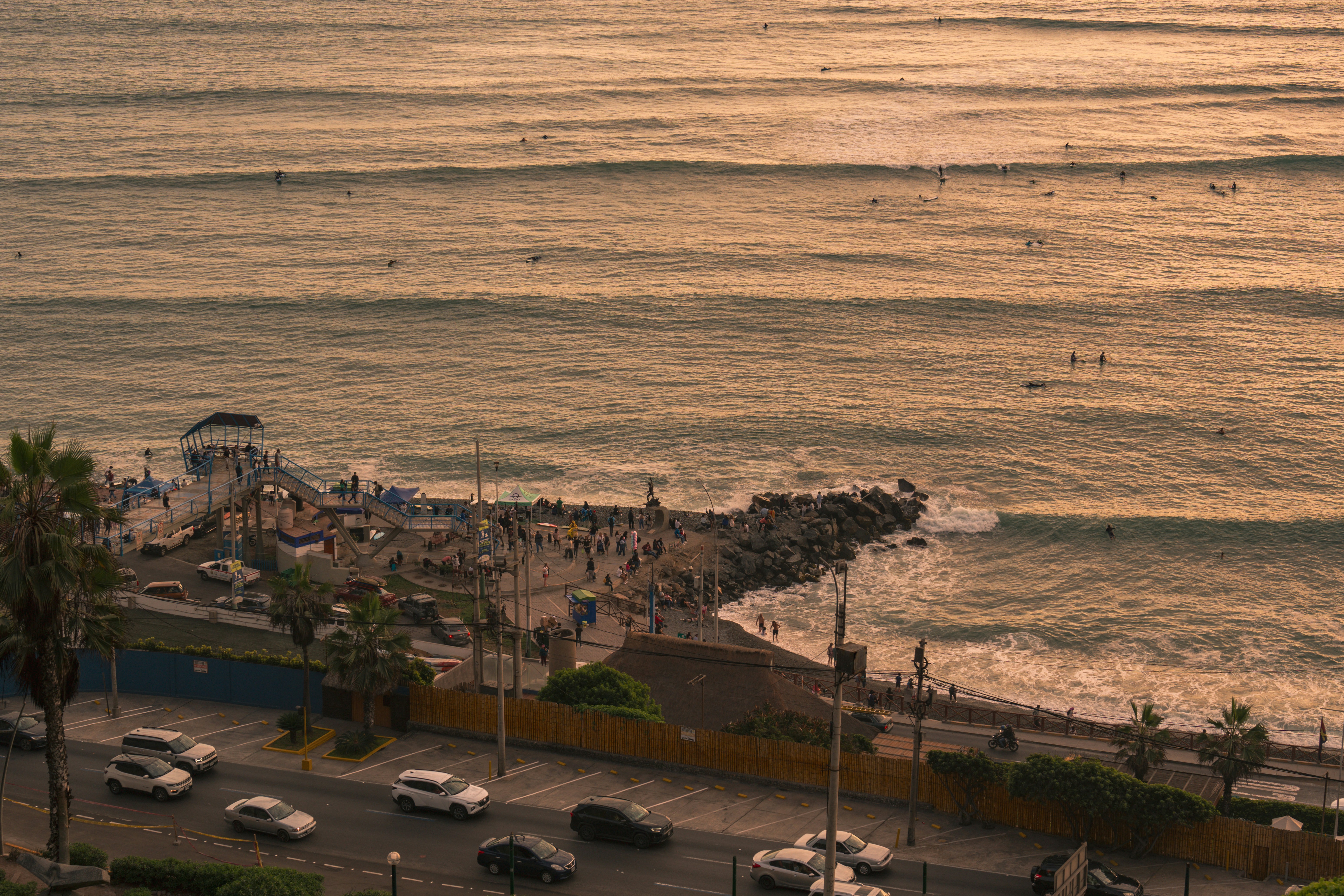 Crowded beach and rocky pier at sunset with waves lapping the shore and cars passing by on the adjacent road.