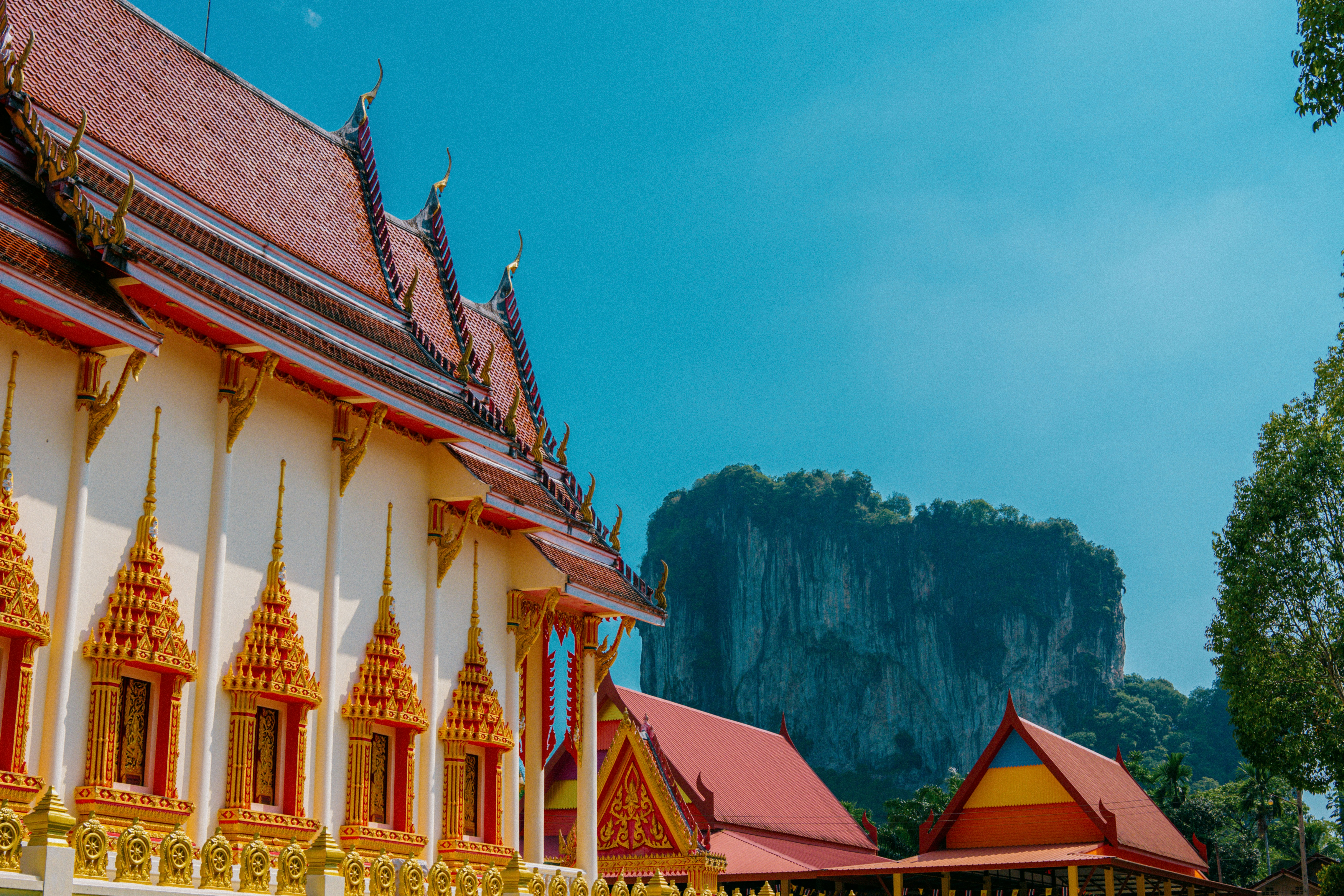Intricate Thai temple architecture juxtaposed against a majestic limestone mountain backdrop under a clear blue sky.