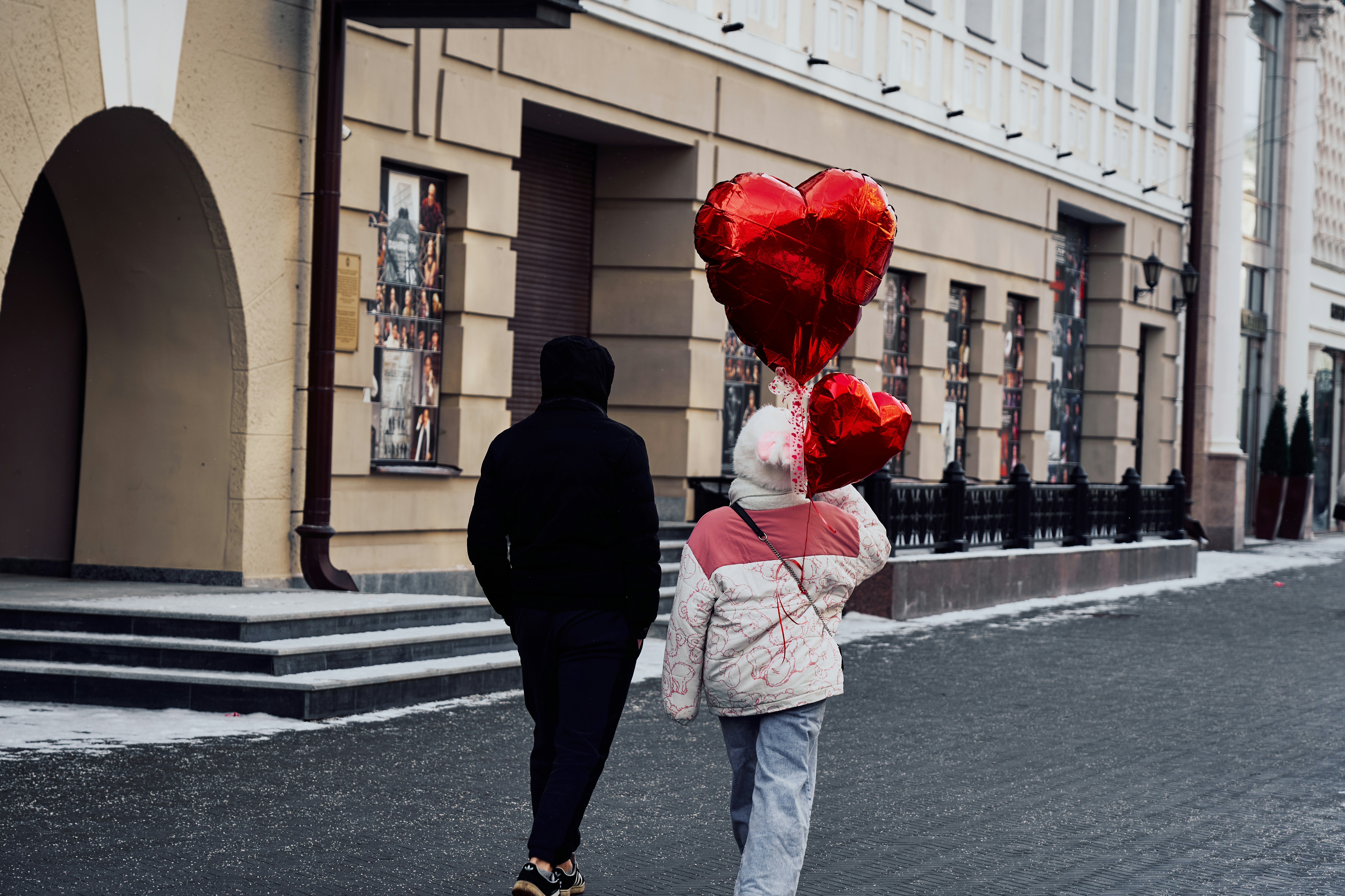 Two wanderers with red heart balloons drift through a quiet cobblestone street