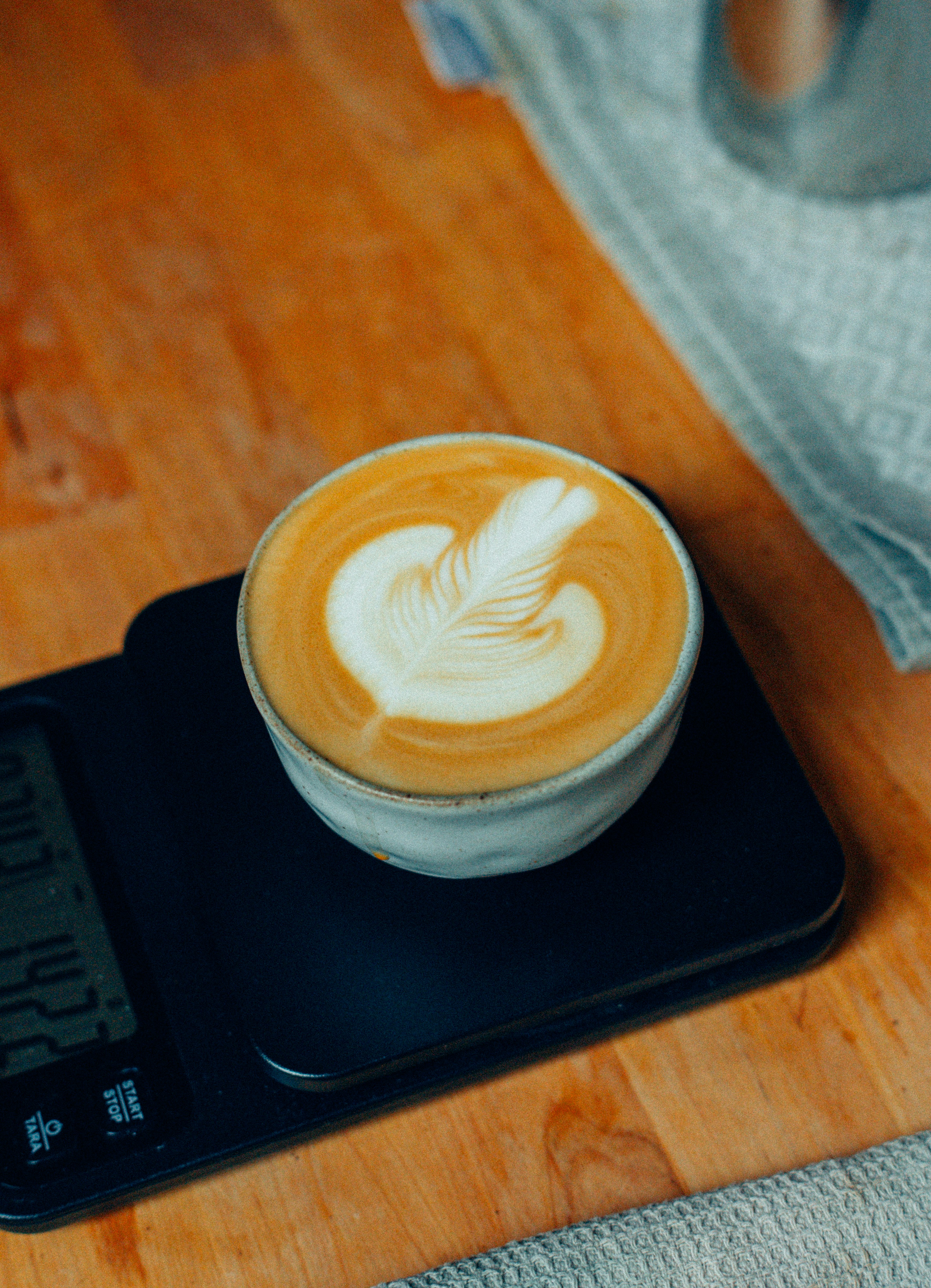 Ceramic cup with latte art heart sits on a black digital scale atop a wooden surface, capturing a quiet cafe-style morning moment.