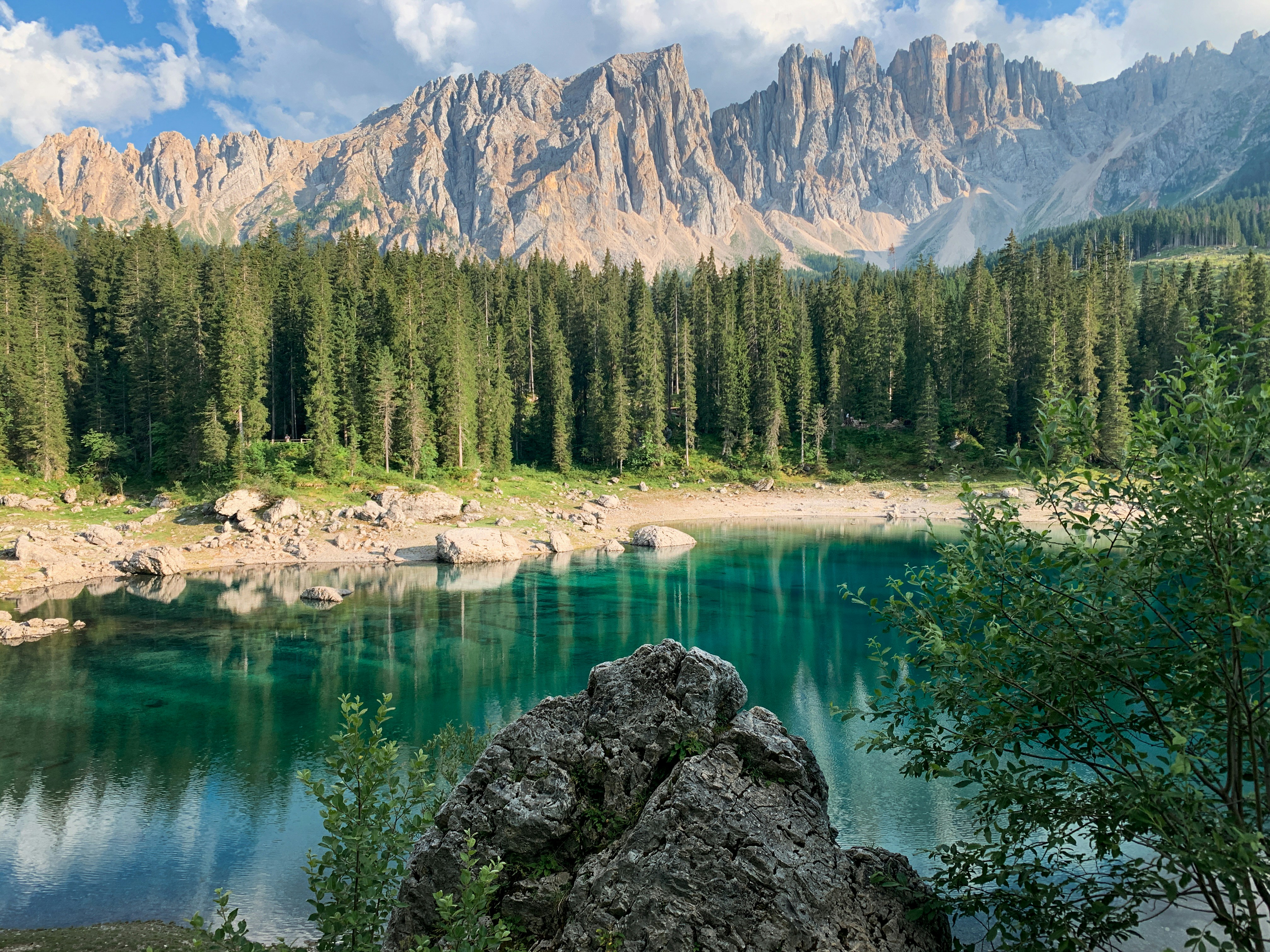 Mountain lake reflecting rocky cliffs and pine forests under a partly cloudy sky.