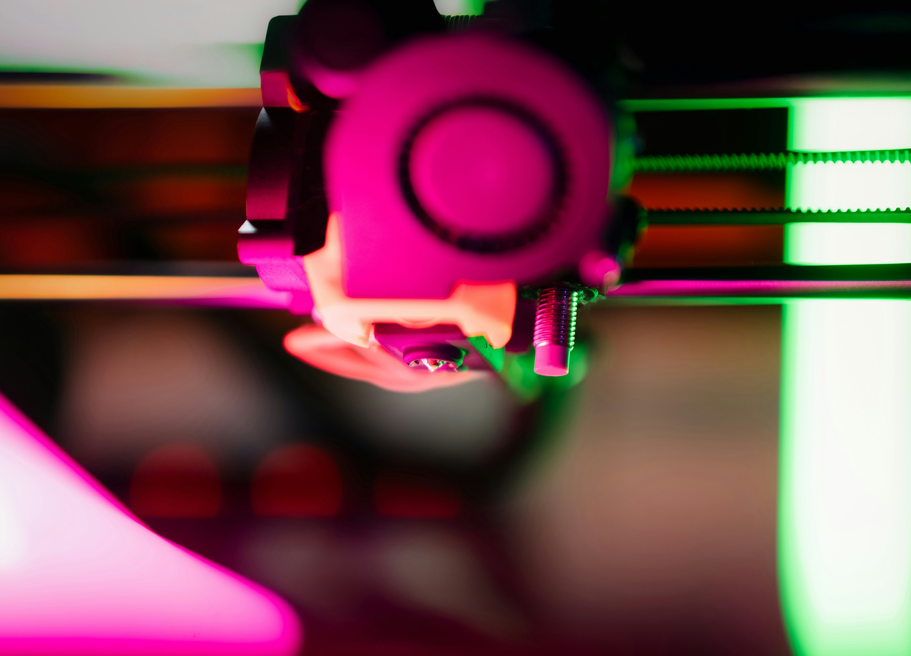 Close-up of a 3D printer nozzle illuminated in vibrant pink and green hues.