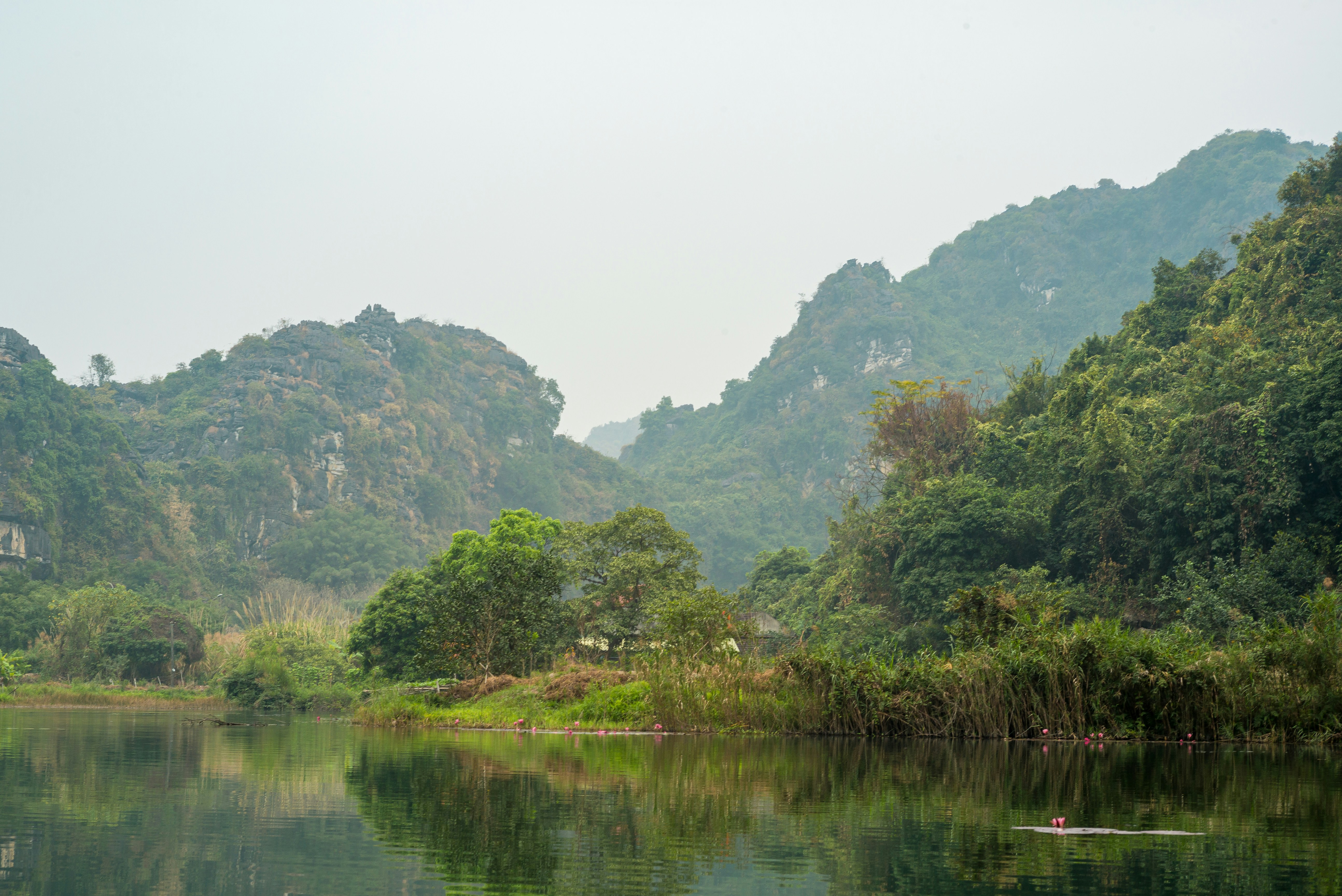 Lush green hills reflecting in a calm, misty lake under an overcast sky.