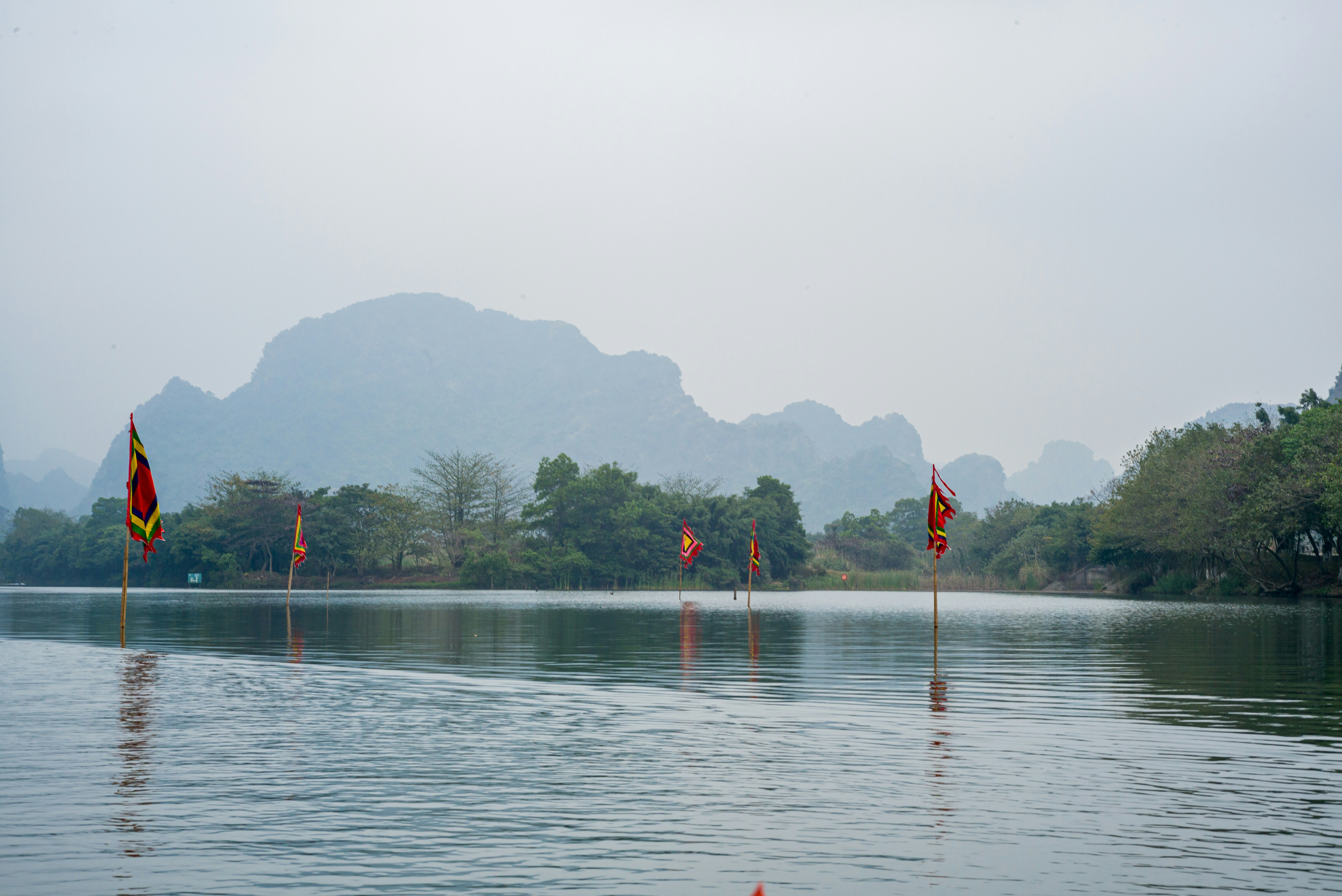 Colorful flags stand in calm water against misty mountains.