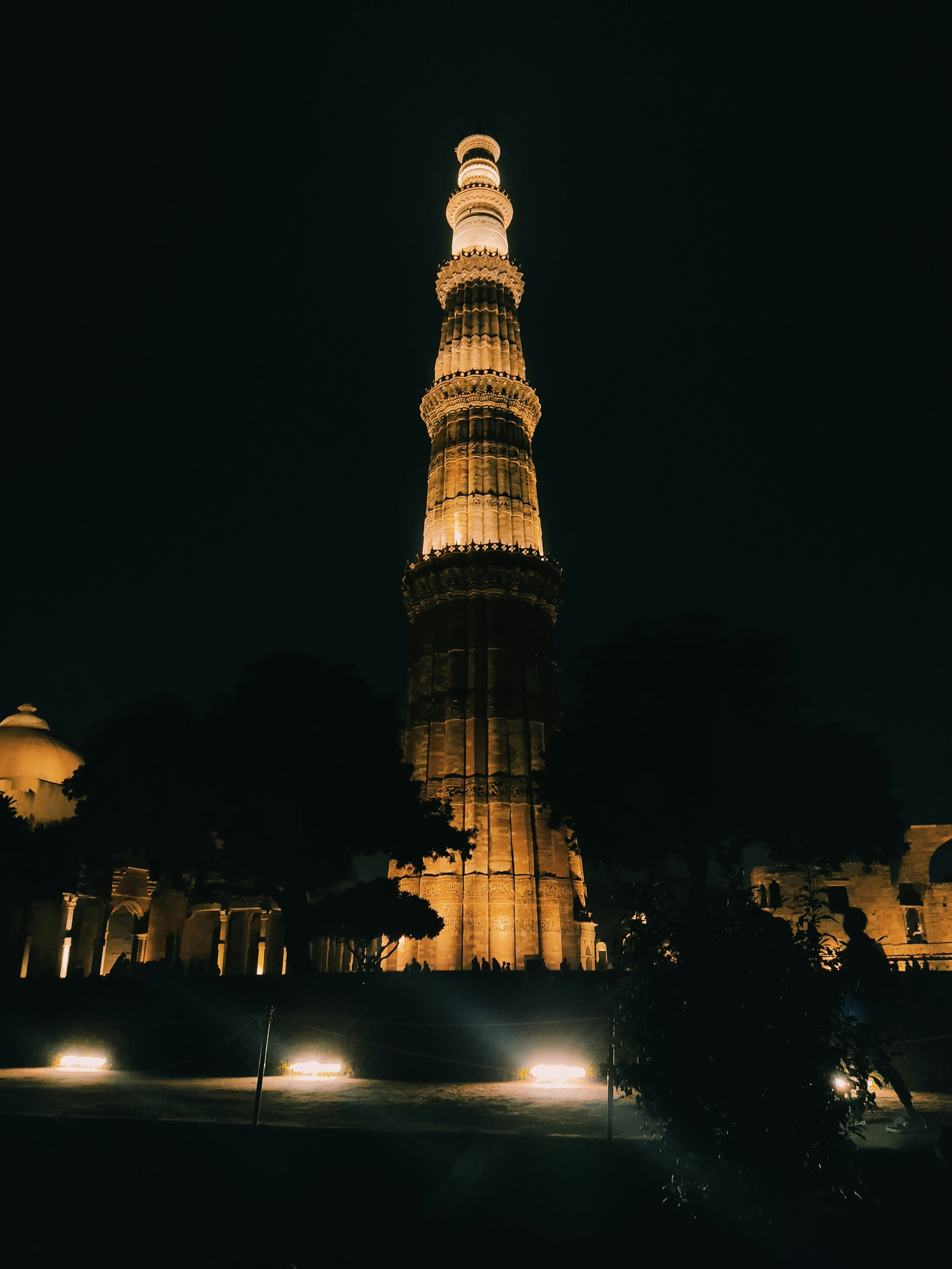 Tall minaret lit in warm amber tones rises above a low skyline, with surrounding silhouettes and ground lights anchoring the scene. Photograph captures a nighttime architectural moment with a focus on the tower’s height and texture.