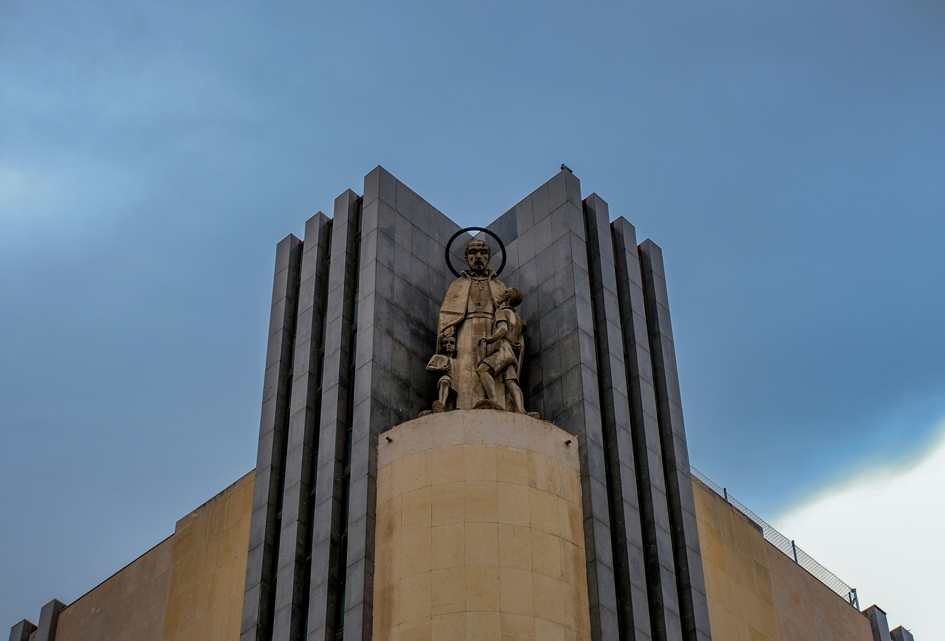 Stone statue atop a modernist building with a dramatic cloudy sky backdrop.