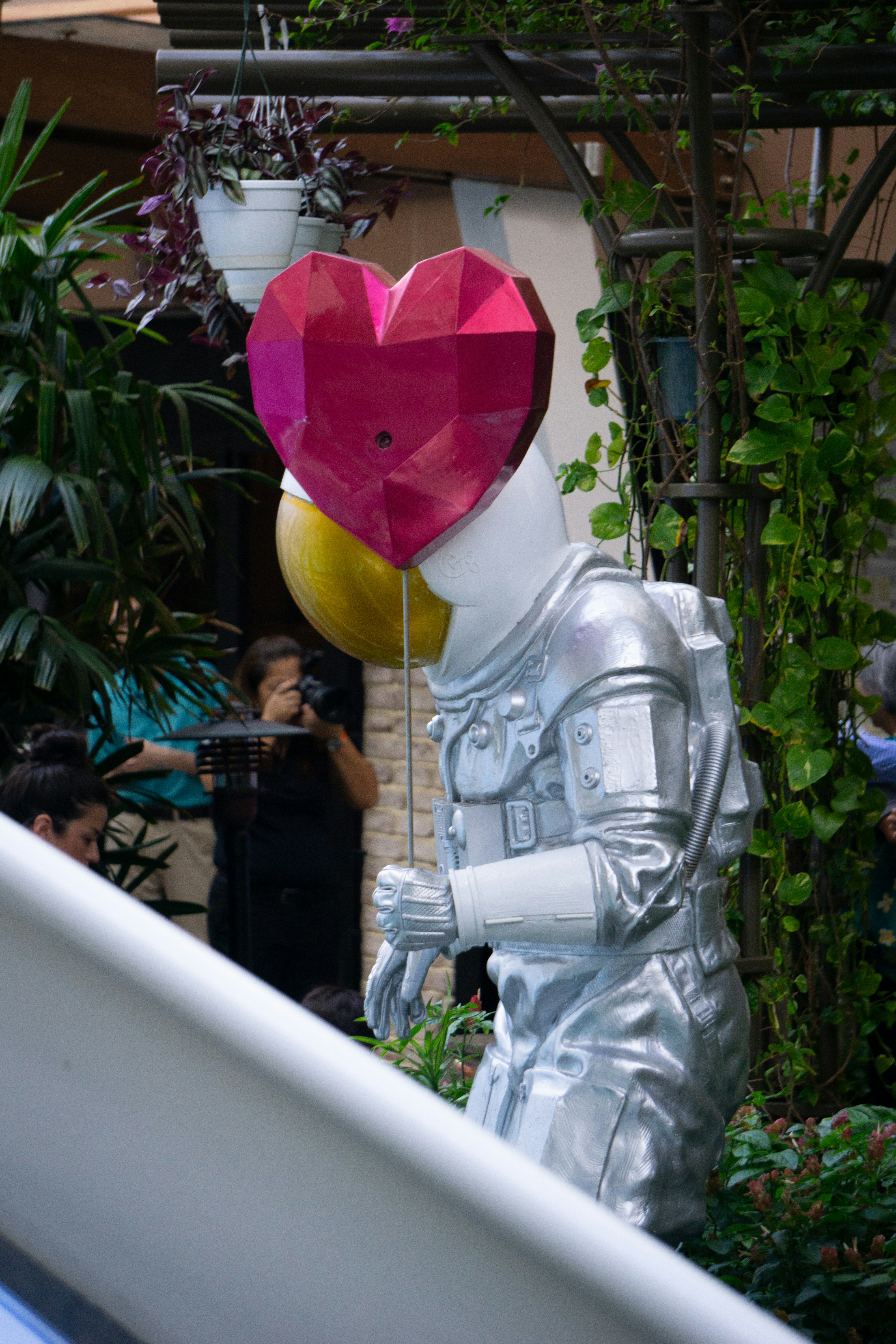 Faceted Heart Helmet on a Silver Astronaut in a Lush Courtyard