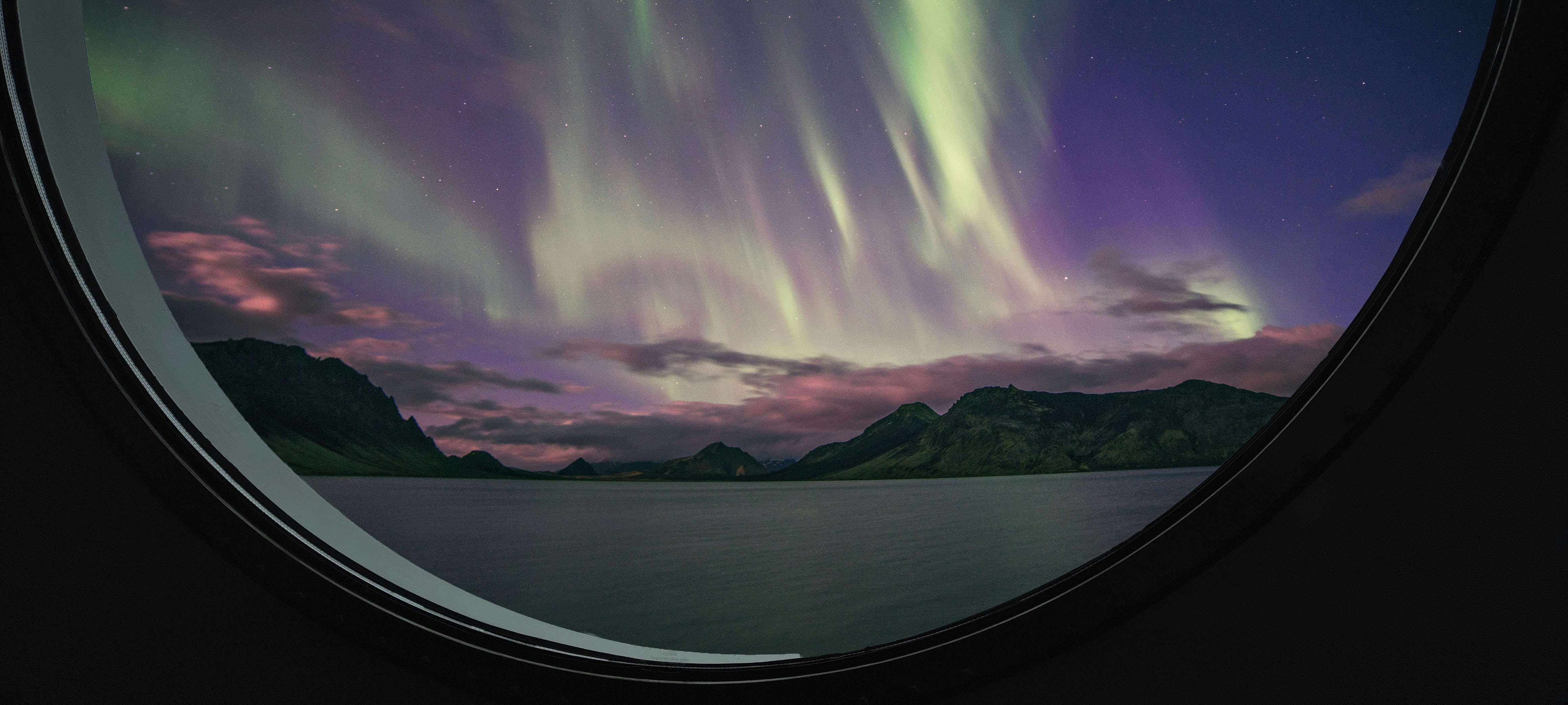 Northern lights dance above a mountainous landscape viewed through a circular window at dusk.