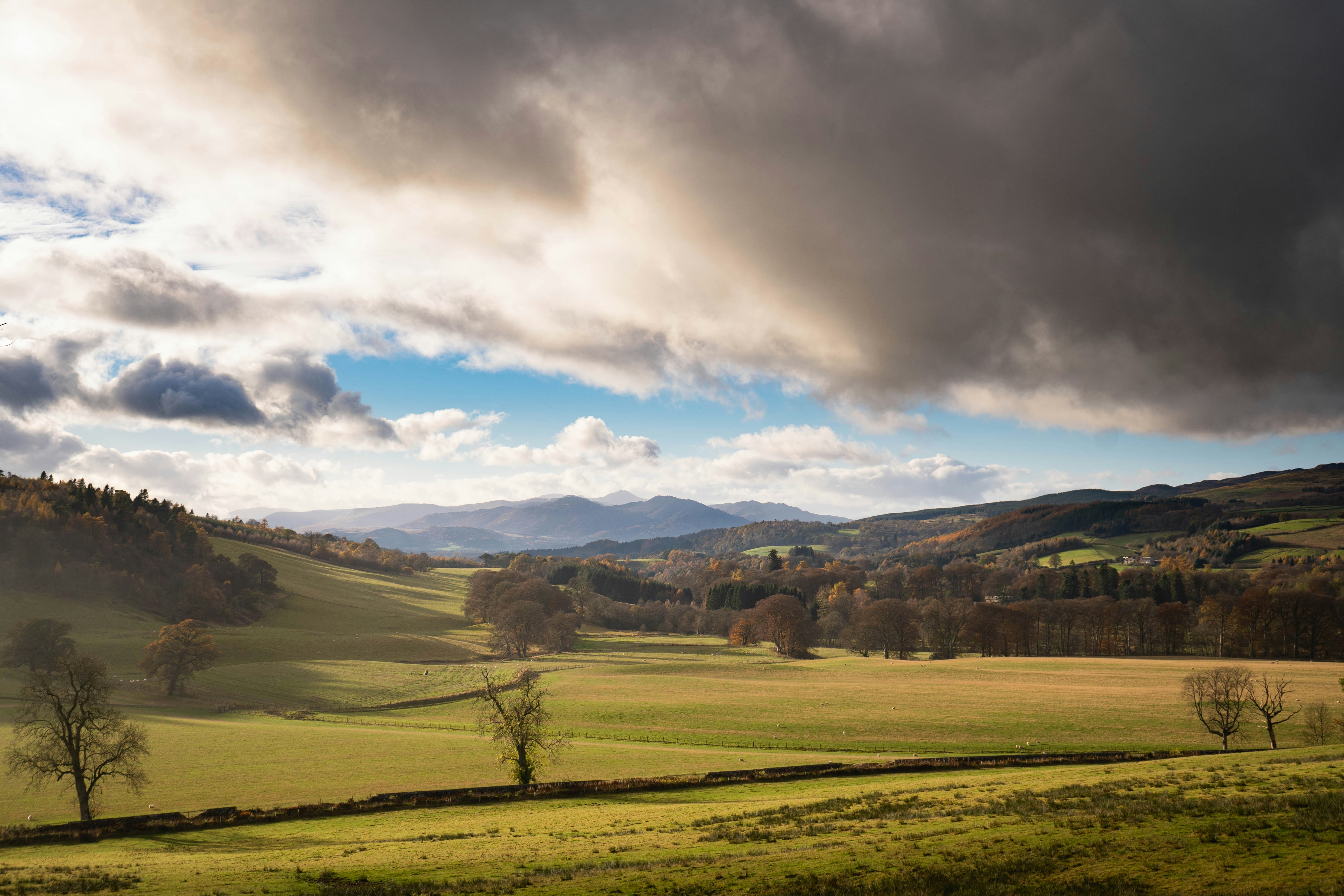 Expansive green landscape under a dynamic sky with dark clouds and distant mountains.
