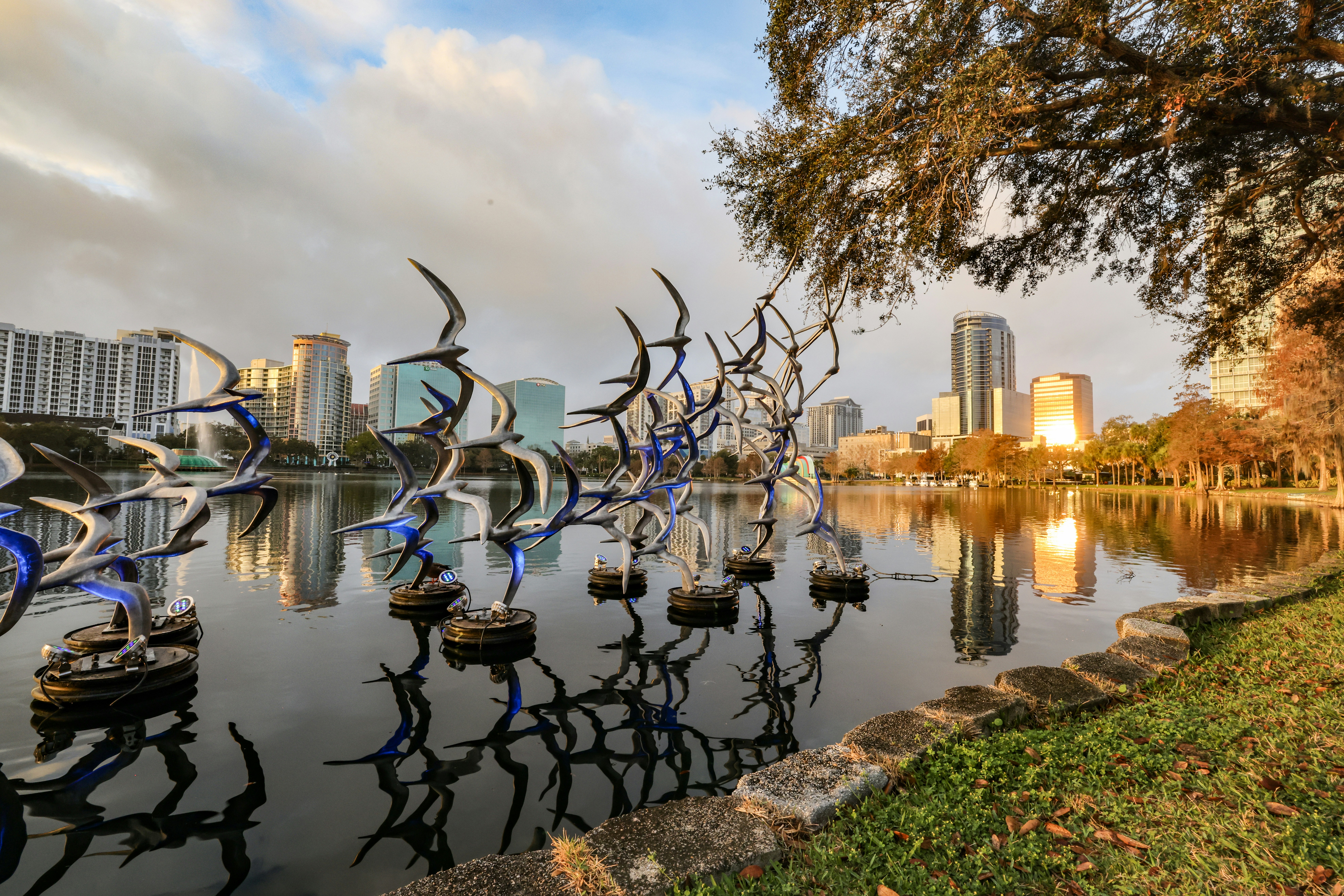 Metallic bird sculptures reflect on a tranquil lake against a city skyline at sunrise in Orlando.