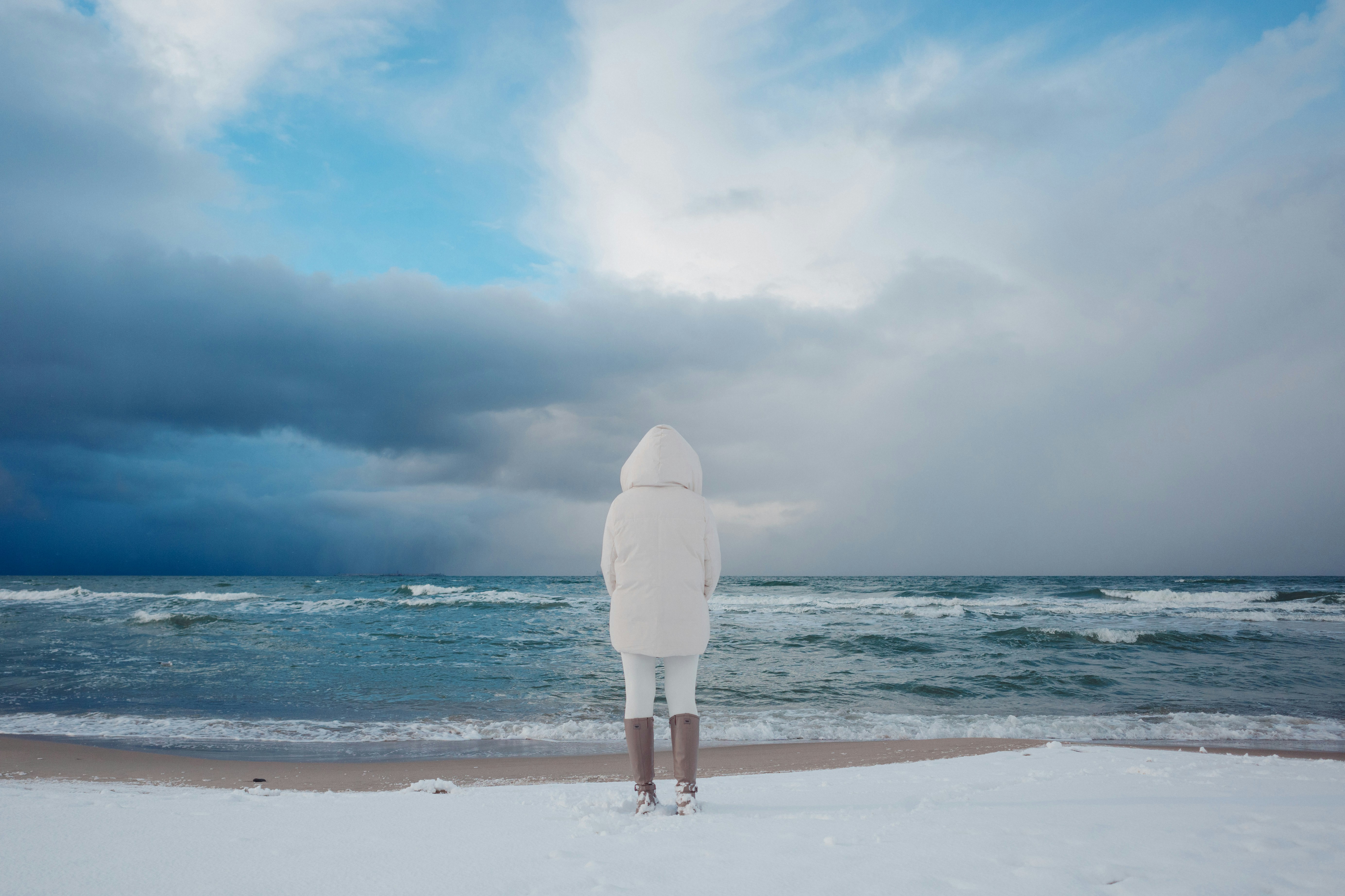 Person in white coat stands on snowy beach facing the ocean under a dramatic cloudy sky.