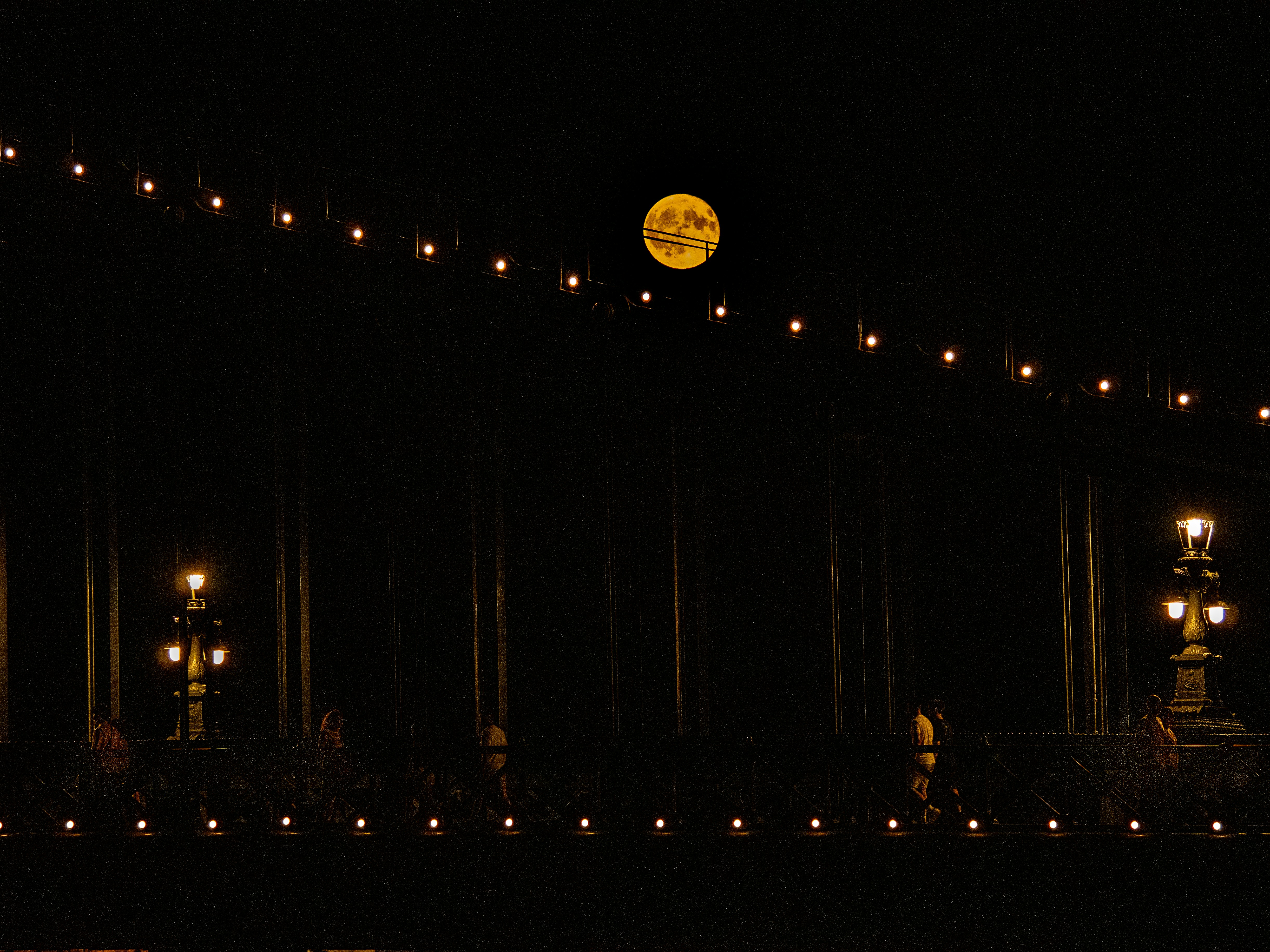Full moon illuminating a bridge with glowing street lamps and silhouettes of pedestrians at night.
