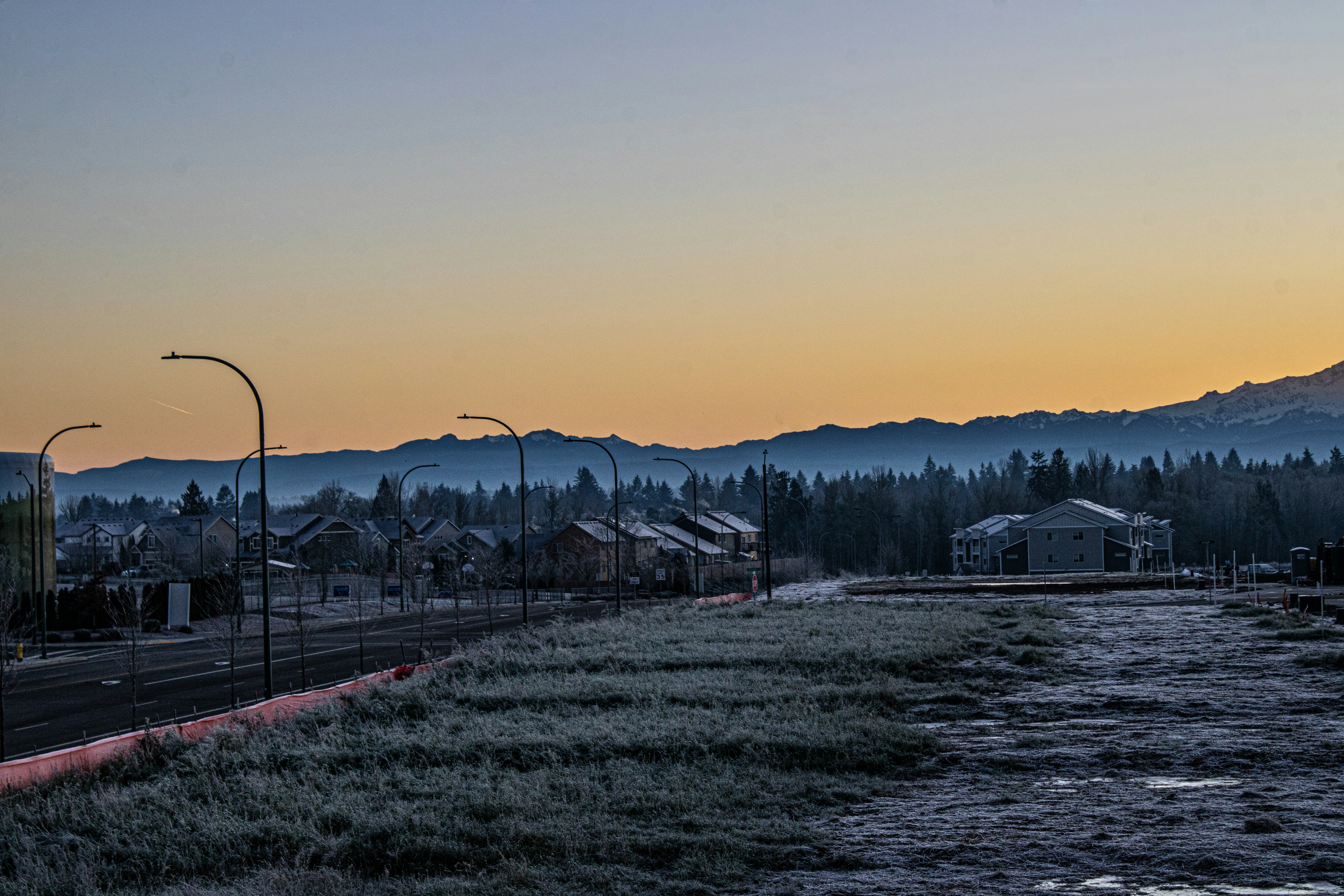 Suburban landscape at sunrise with frost-covered grass and distant mountains under a warm orange sky.