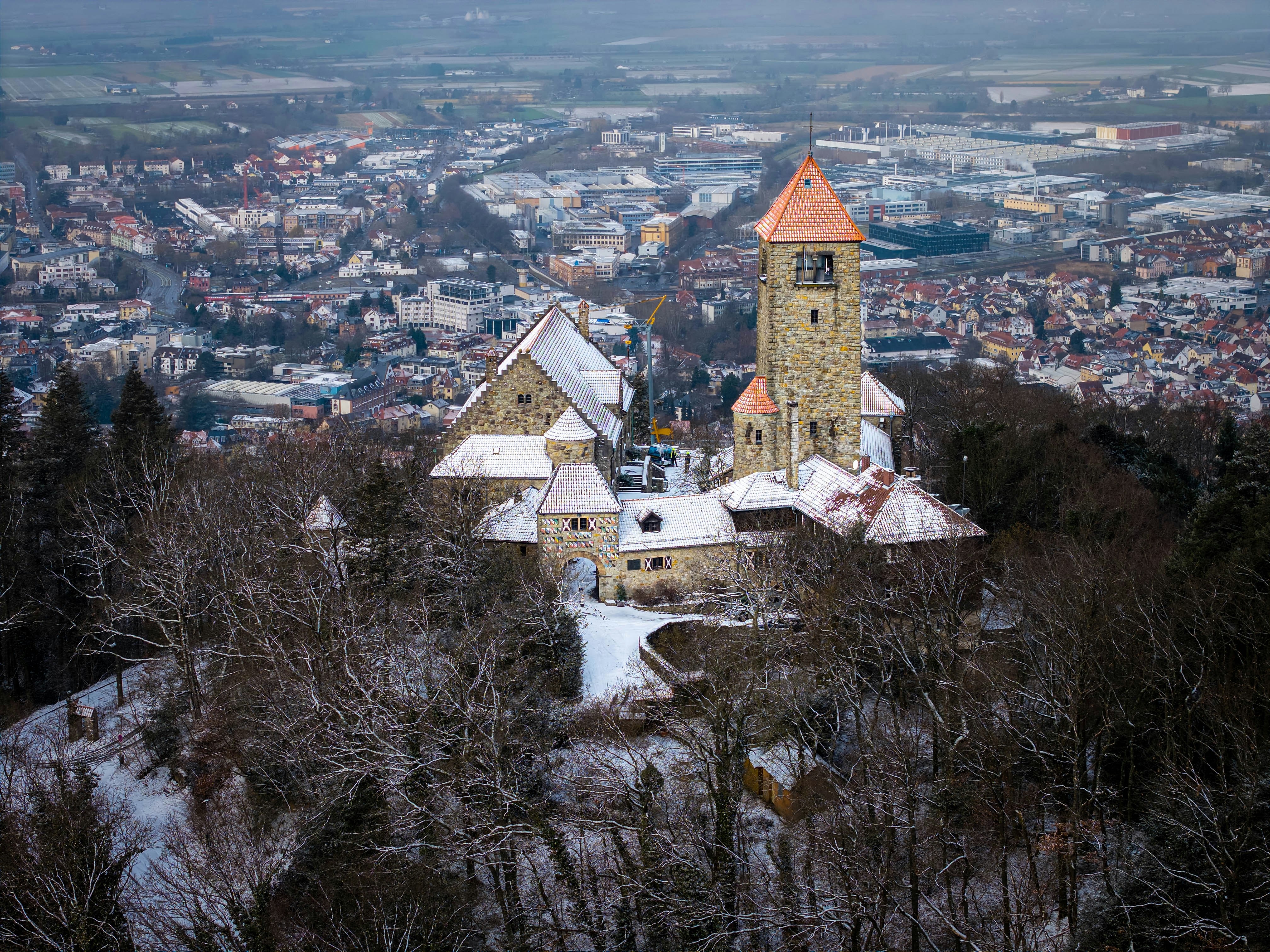 Snow-dusted castle amidst bare winter trees with an expansive urban backdrop.