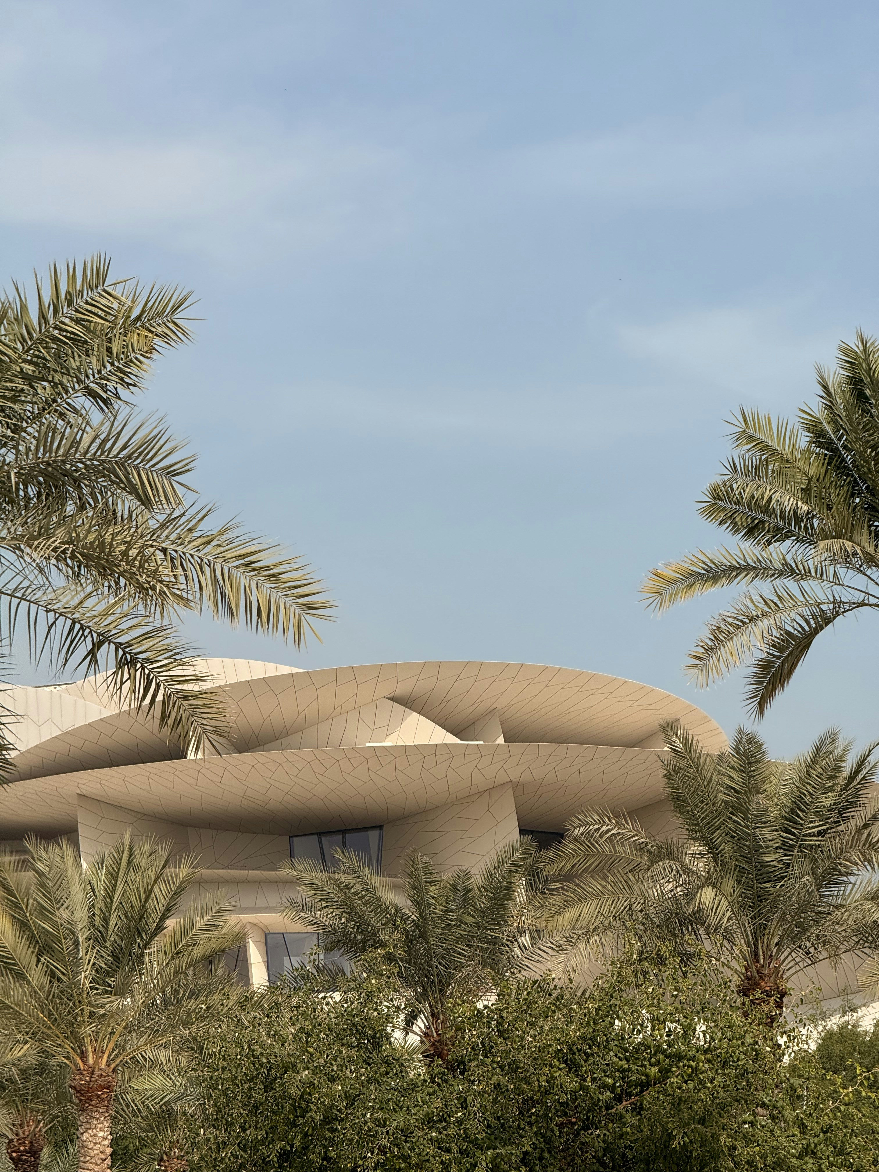 Photograph of modern, shell-like architecture rising behind a cluster of palms under a clear blue sky.