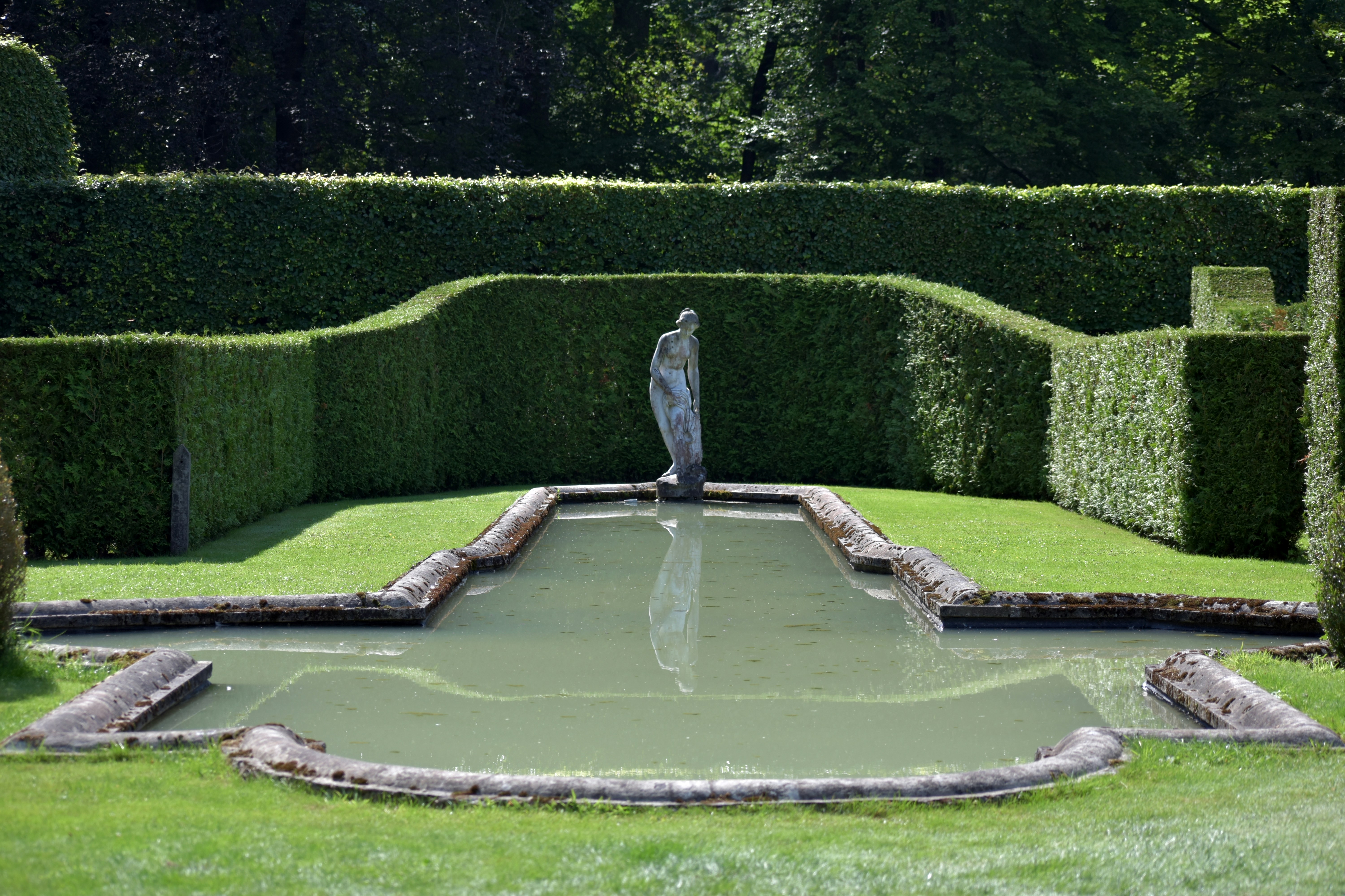 Stone statue overlooks a serene pond surrounded by manicured hedges.