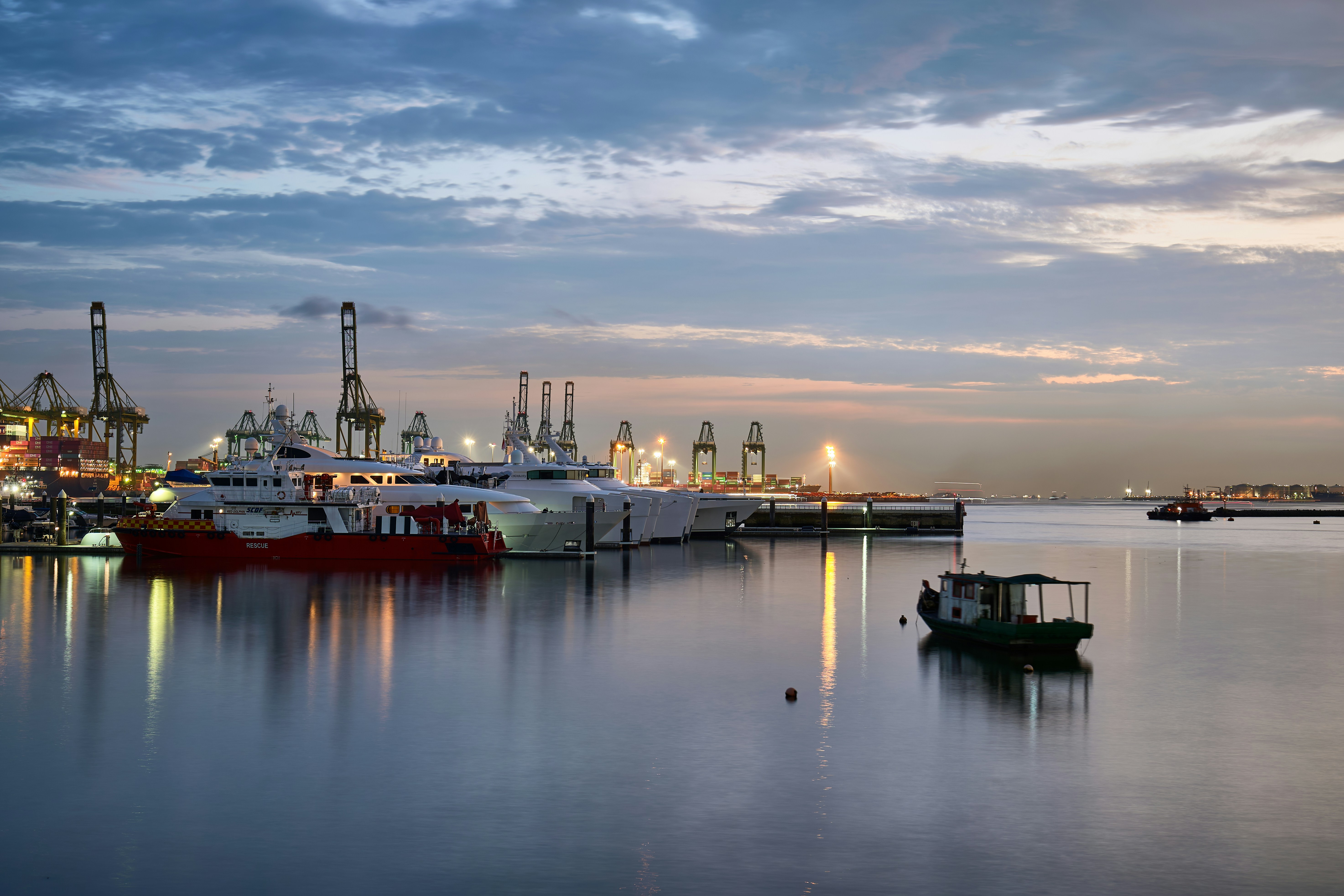 Ships docked at a tranquil harbor under a twilight sky, with industrial cranes silhouetted against the horizon.