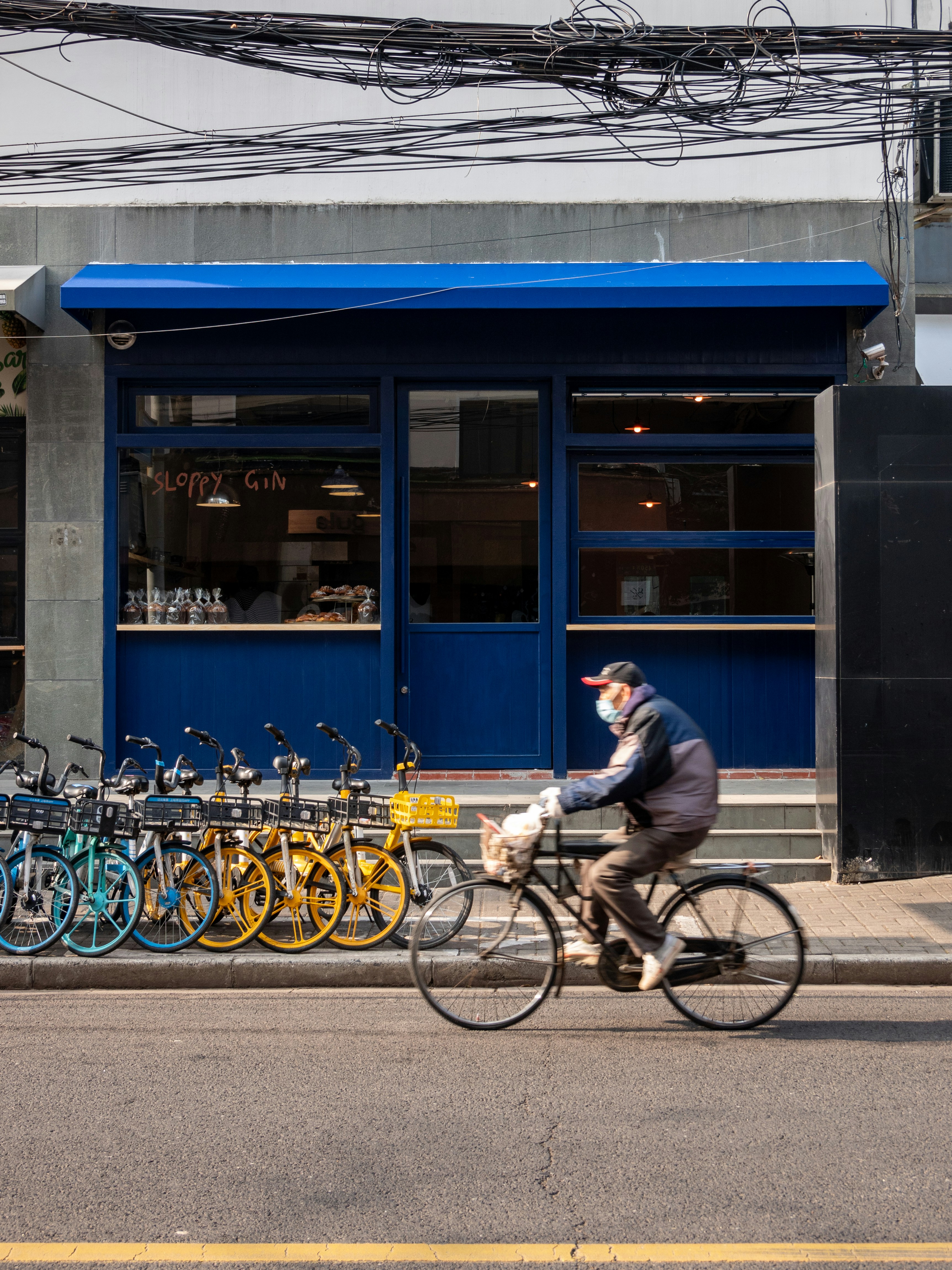 Blue storefront, row of colorful bikes, and a masked cyclist gliding by