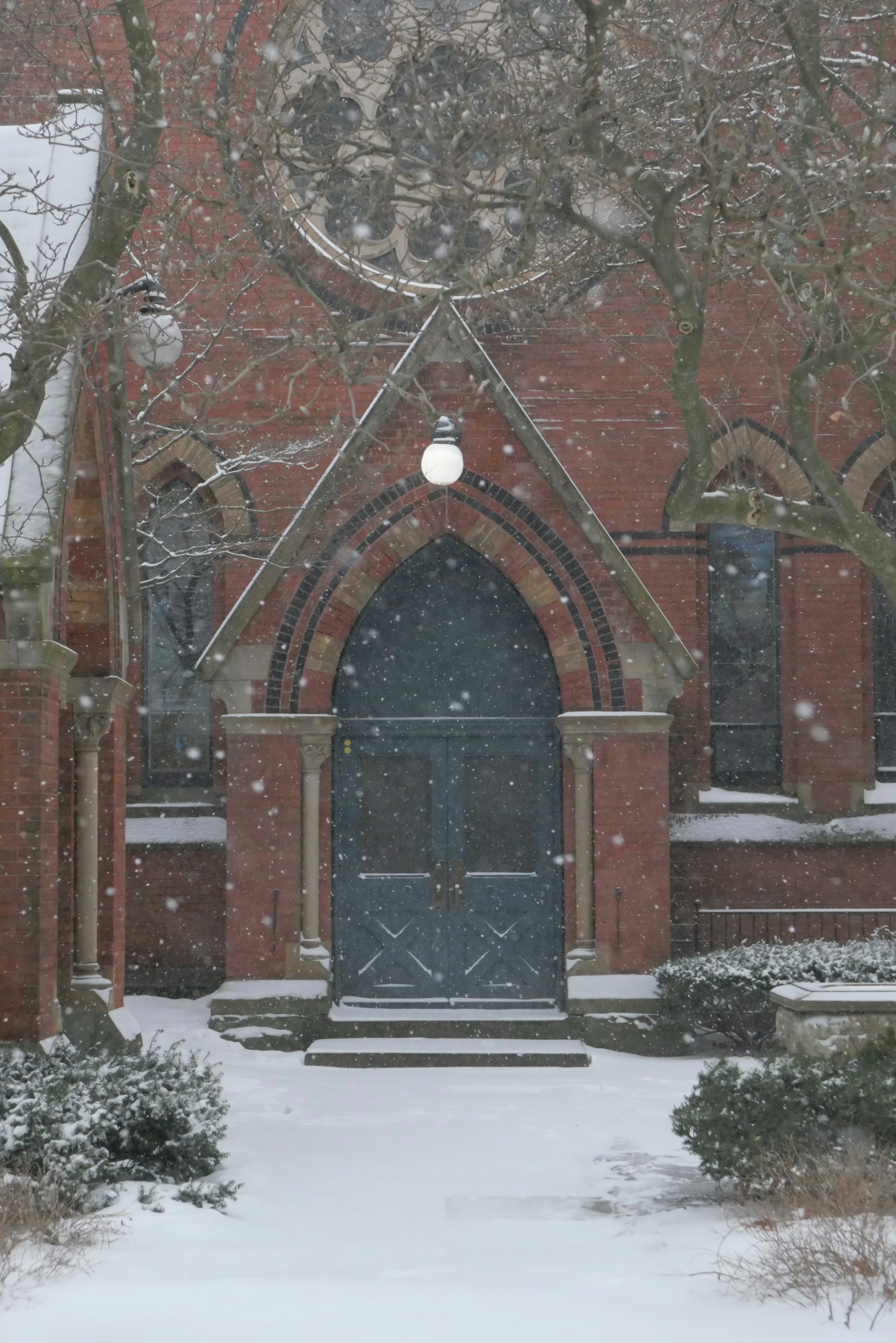 Snow blankets a brick Gothic church entrance framed by trees, with a blue arched door and a circular window above. The scene conveys quiet, wintry stillness.