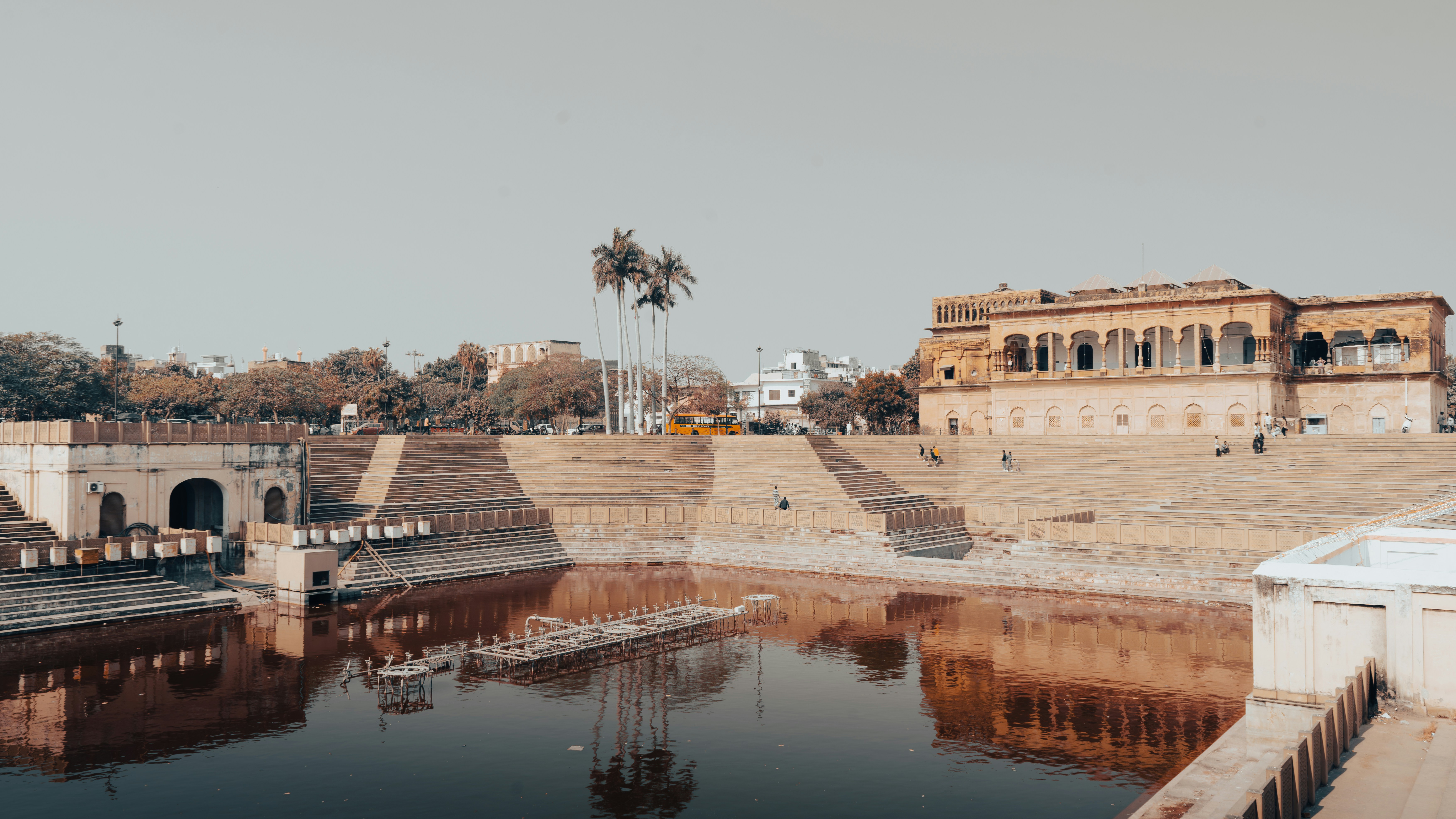 Historic stepwell with arched architecture and palm trees reflected in calm water under a clear sky.