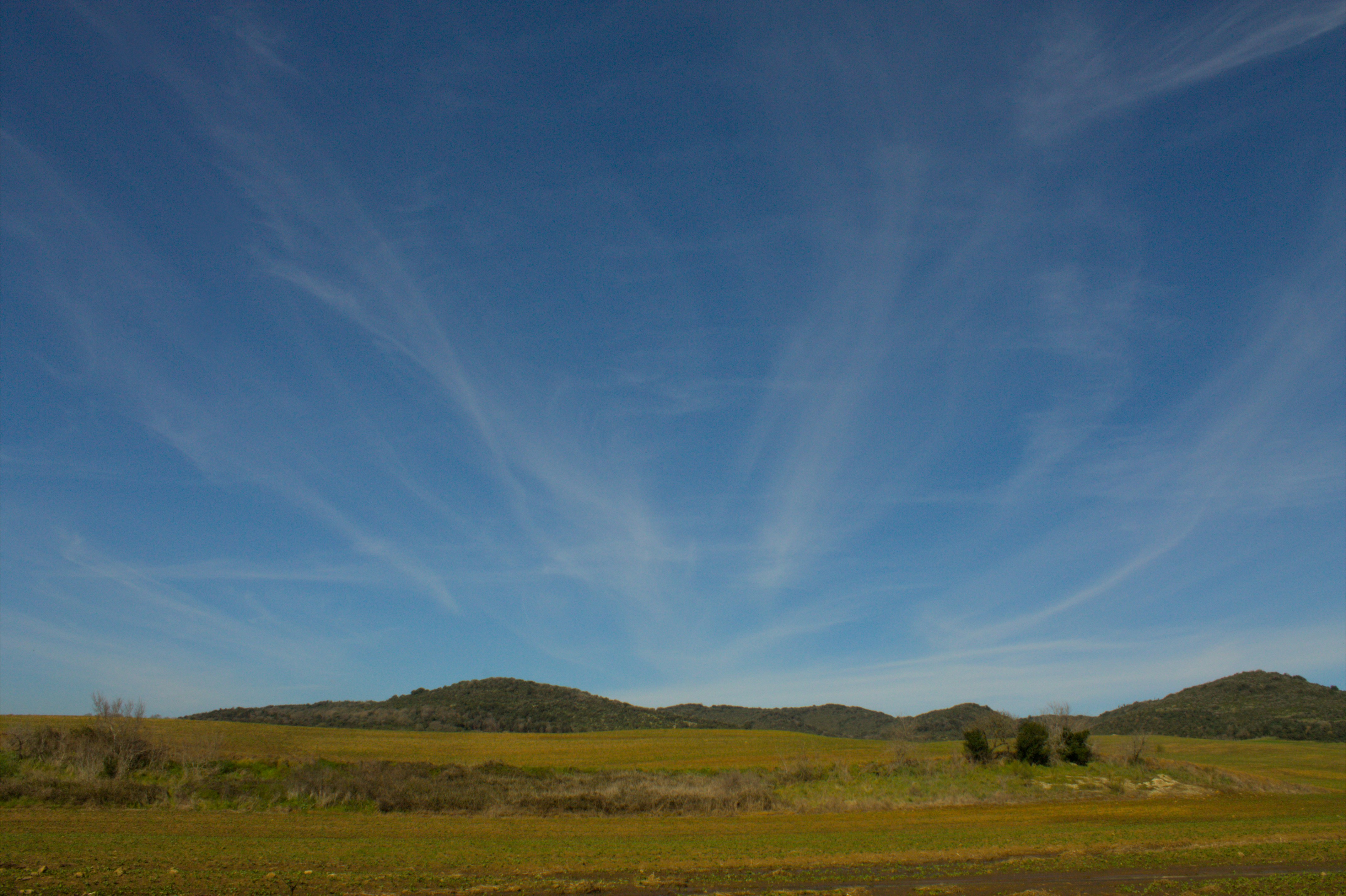 Wispy clouds fan out across a bright blue sky above a serene green landscape with distant hills.