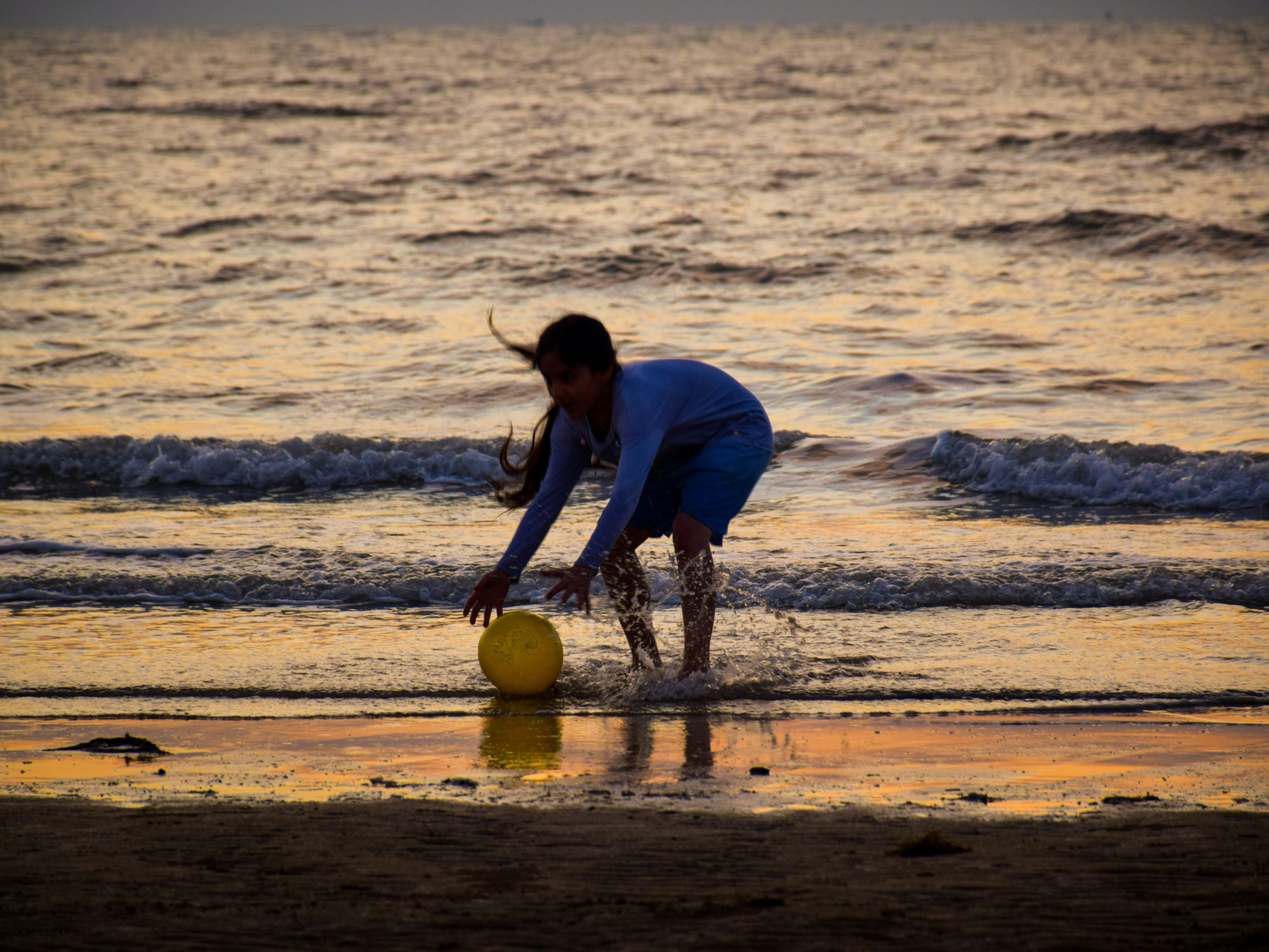 Child in blue attire playing with a yellow ball by the ocean shore at sunset.