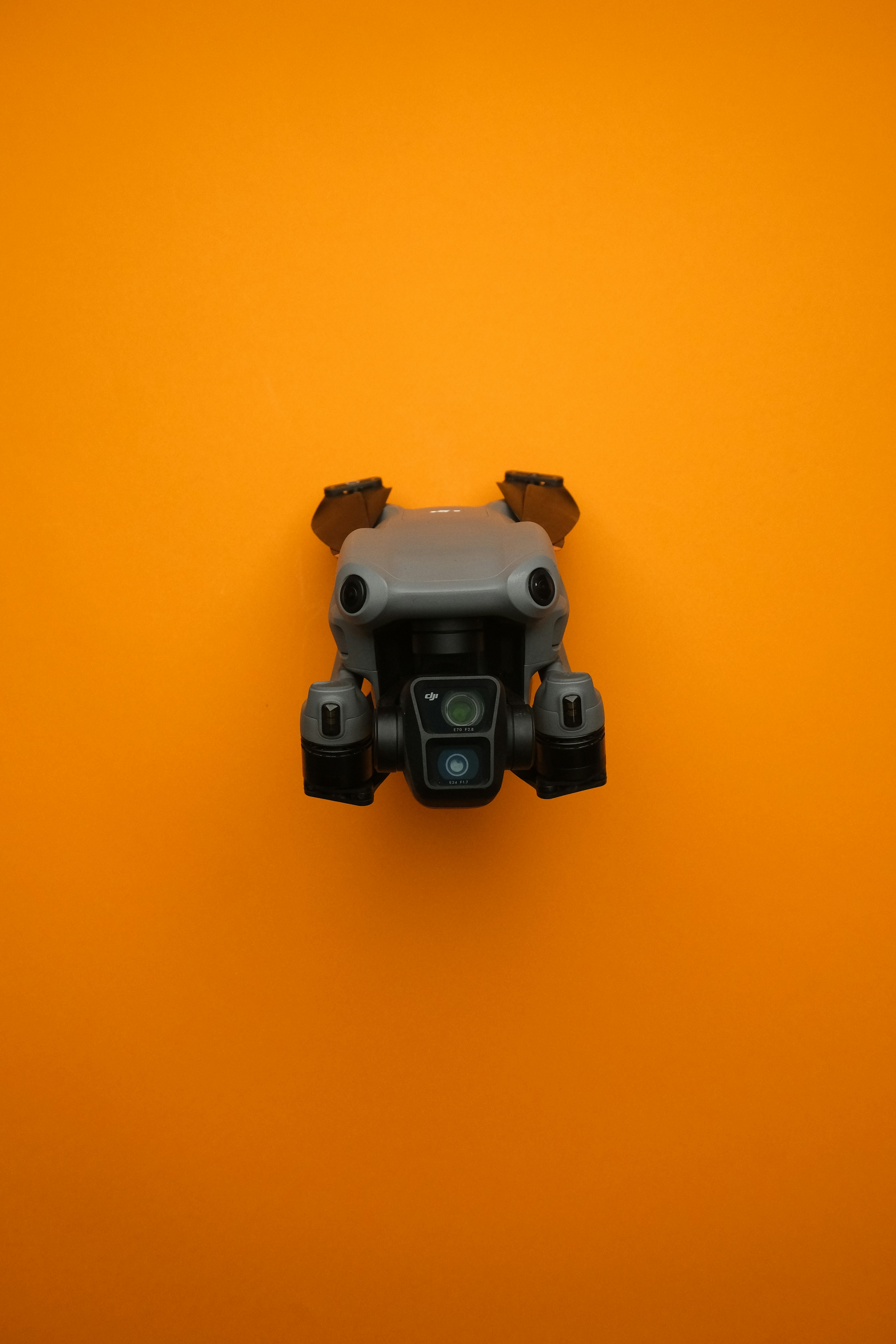 A DJI drone with its front camera and obstacle sensors is placed on a vibrant orange background, showcasing its advanced design and high-tech capabilities. The drone's sleek gray body, dual-lens camera system, and partially unfolded arms highlight its professional build for aerial photography and videography. The contrasting background enhances the modern aesthetic, making this image a perfect representation of cutting-edge drone technology.