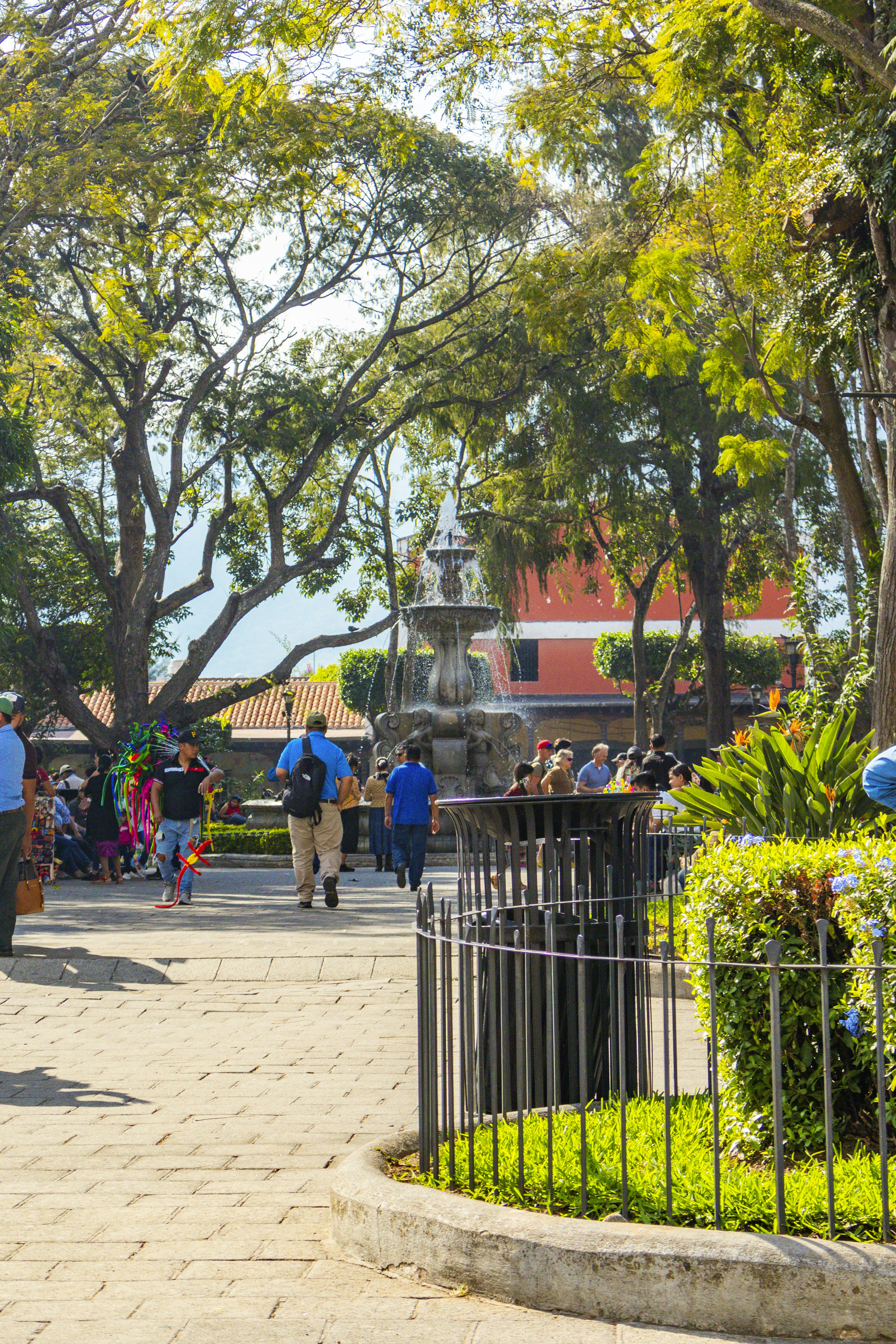 Lively park scene featuring a central fountain surrounded by visitors, lush greenery, and colorful decorations. The atmosphere is filled with joy and community spirit.