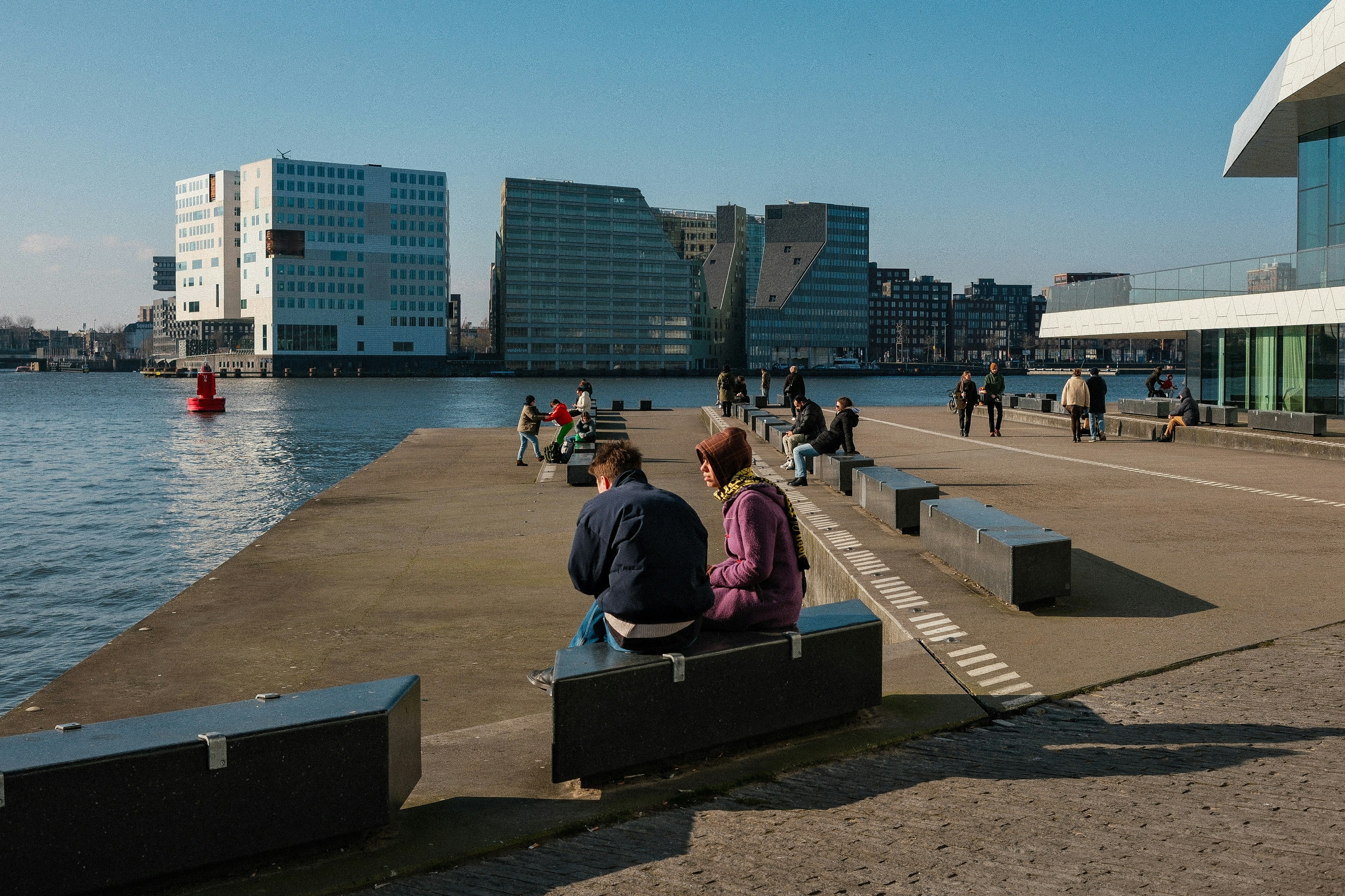 People relaxing on a concrete pier with modern buildings and a clear blue sky in the background.