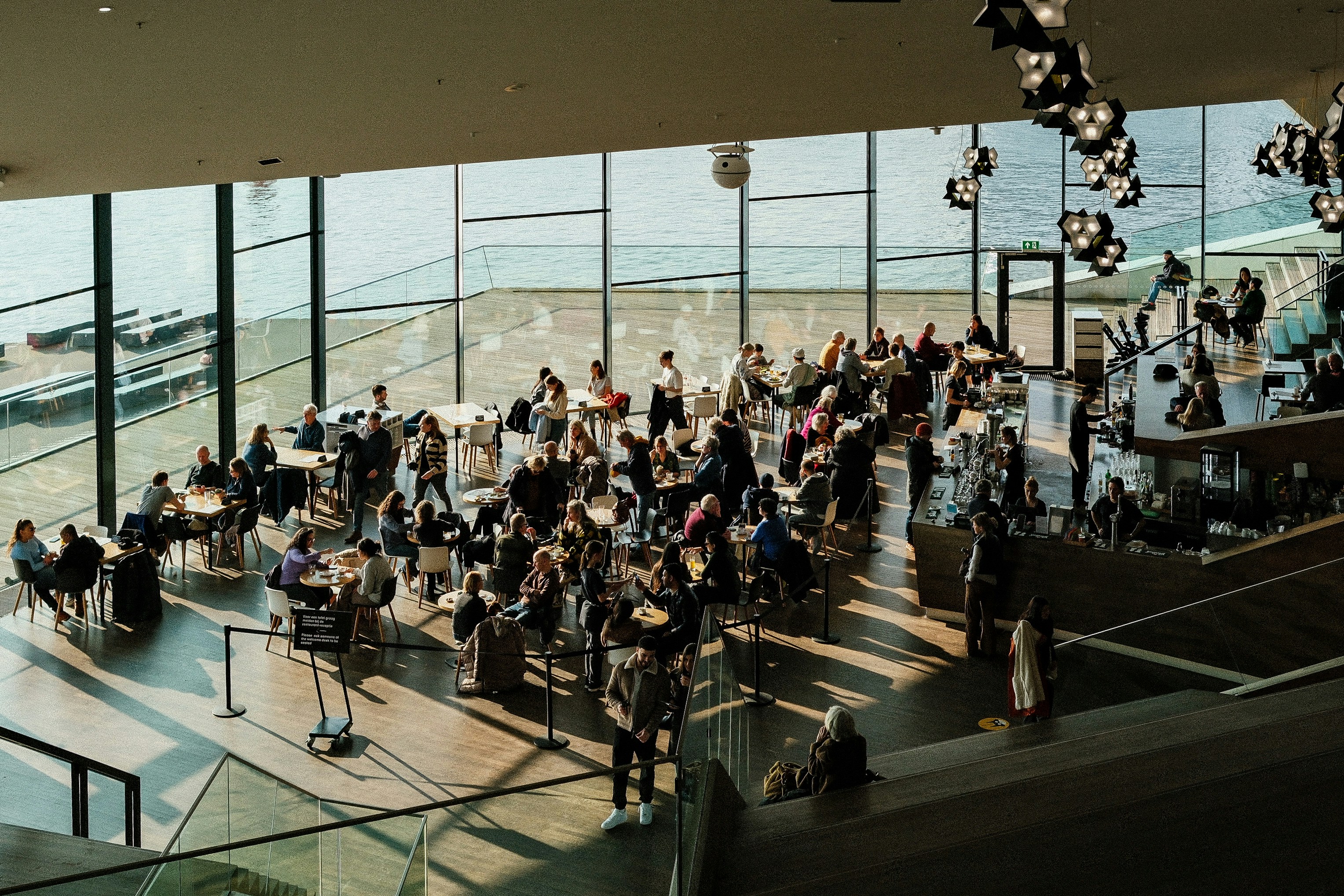 Café patrons enjoying leisurely discussions in a sunlit, modern interior with large windows overlooking the water.