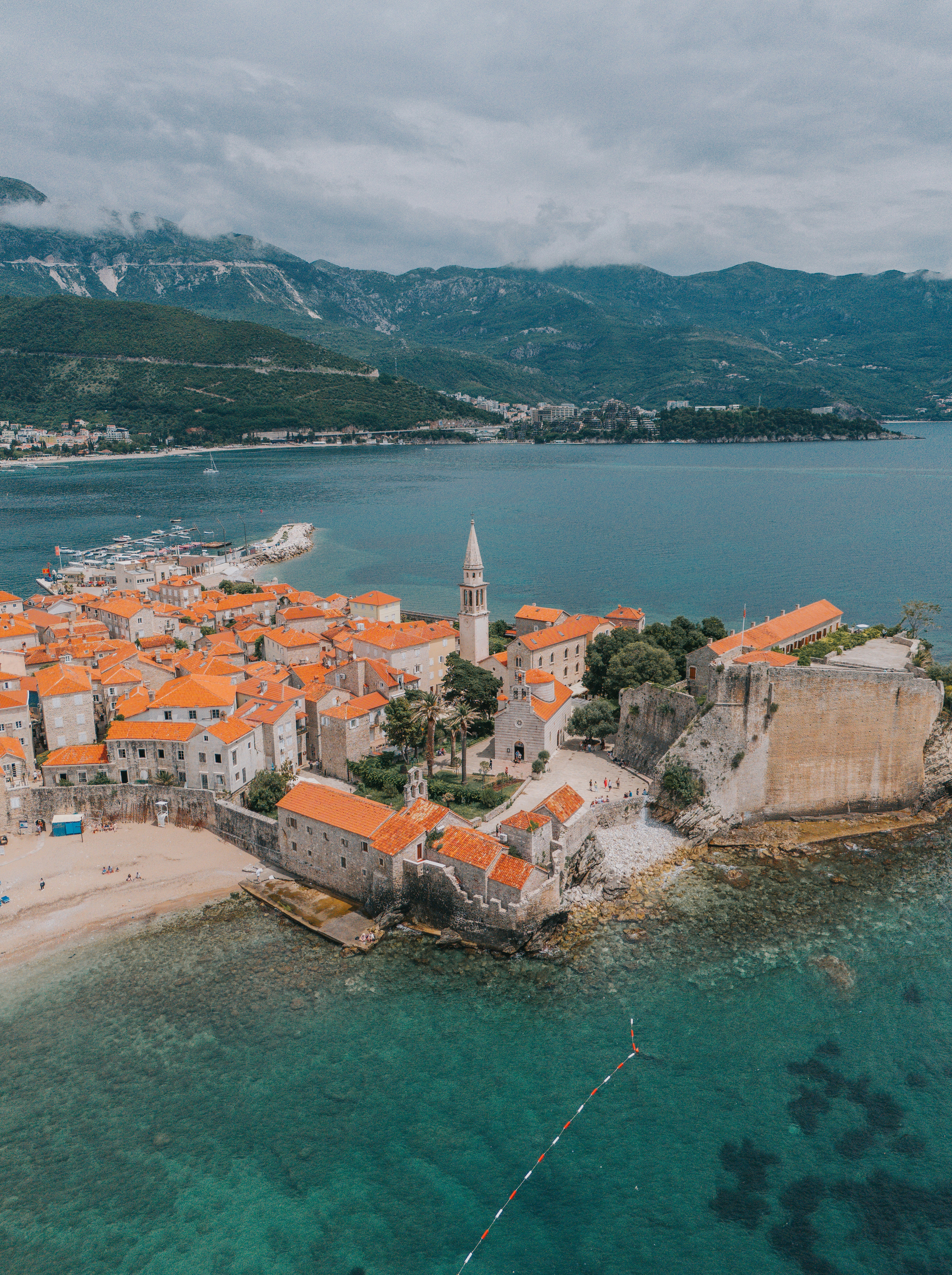Coastal town with orange-tiled roofs and historic architecture set against a turquoise sea and mountainous backdrop.