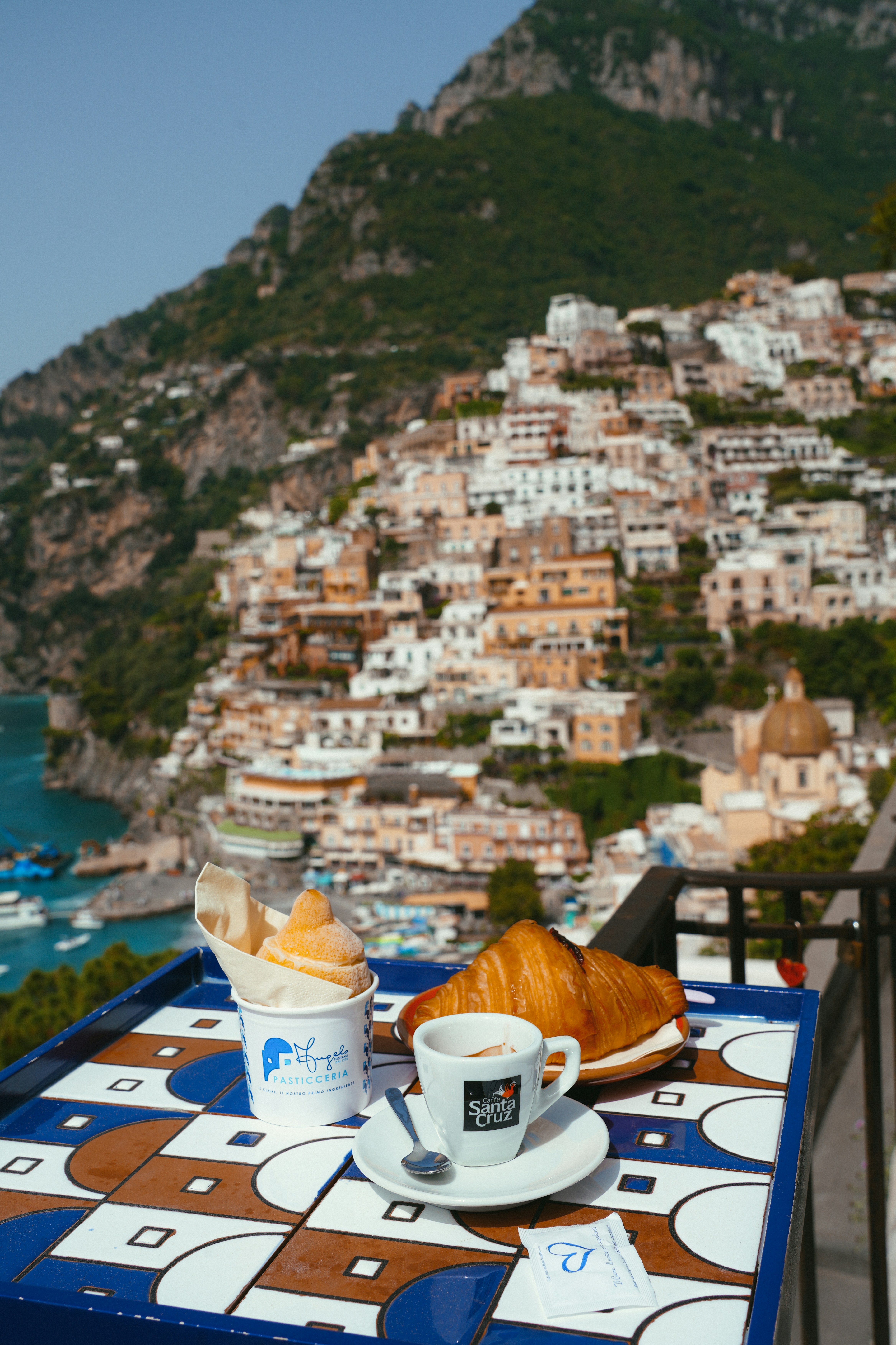 Coffee and croissants on a patterned table with a view of a colorful hillside town by the sea.